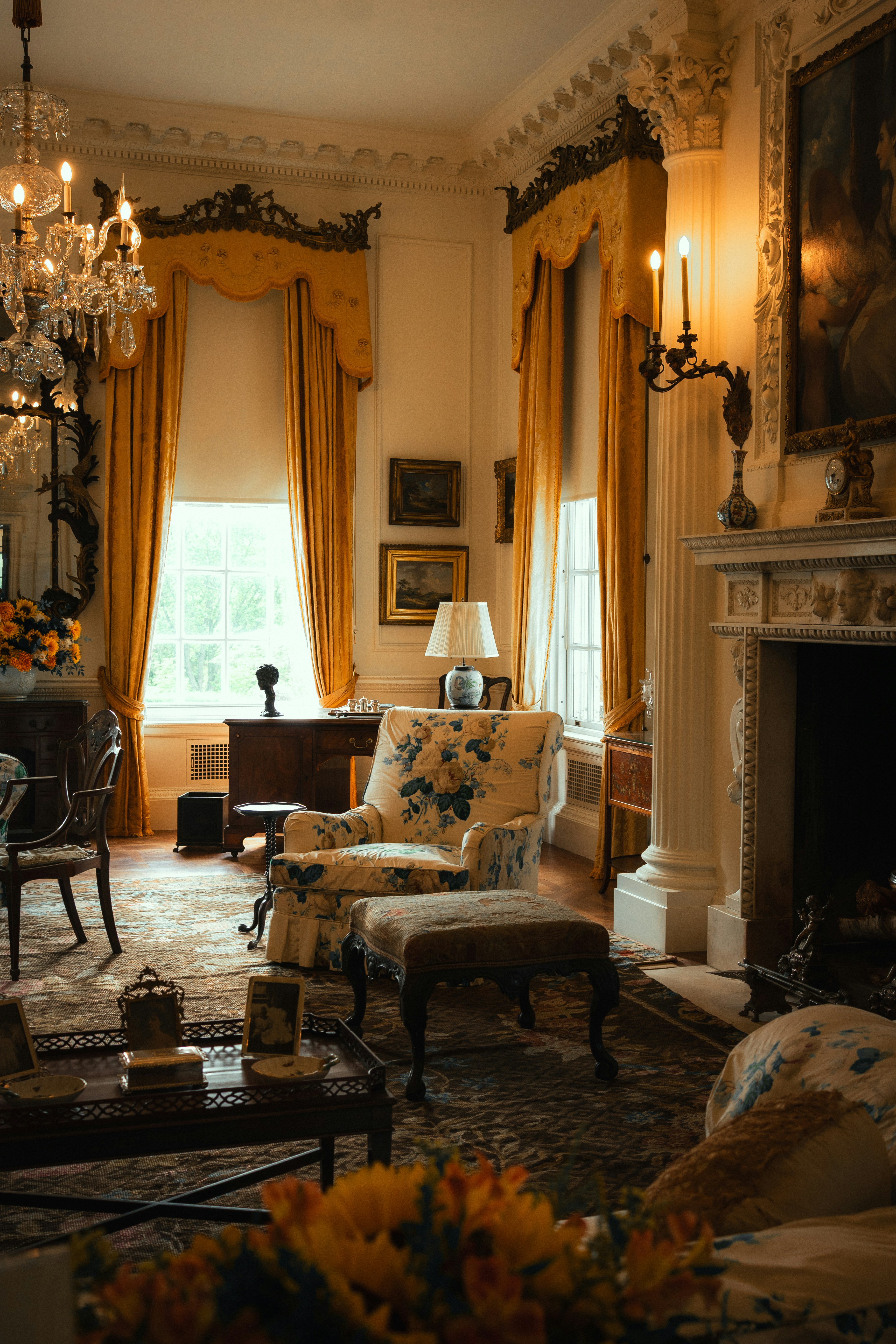 Opulent drawing room with golden drapes, ornate fireplace, and a floral armchair set centered by warm ambient light.