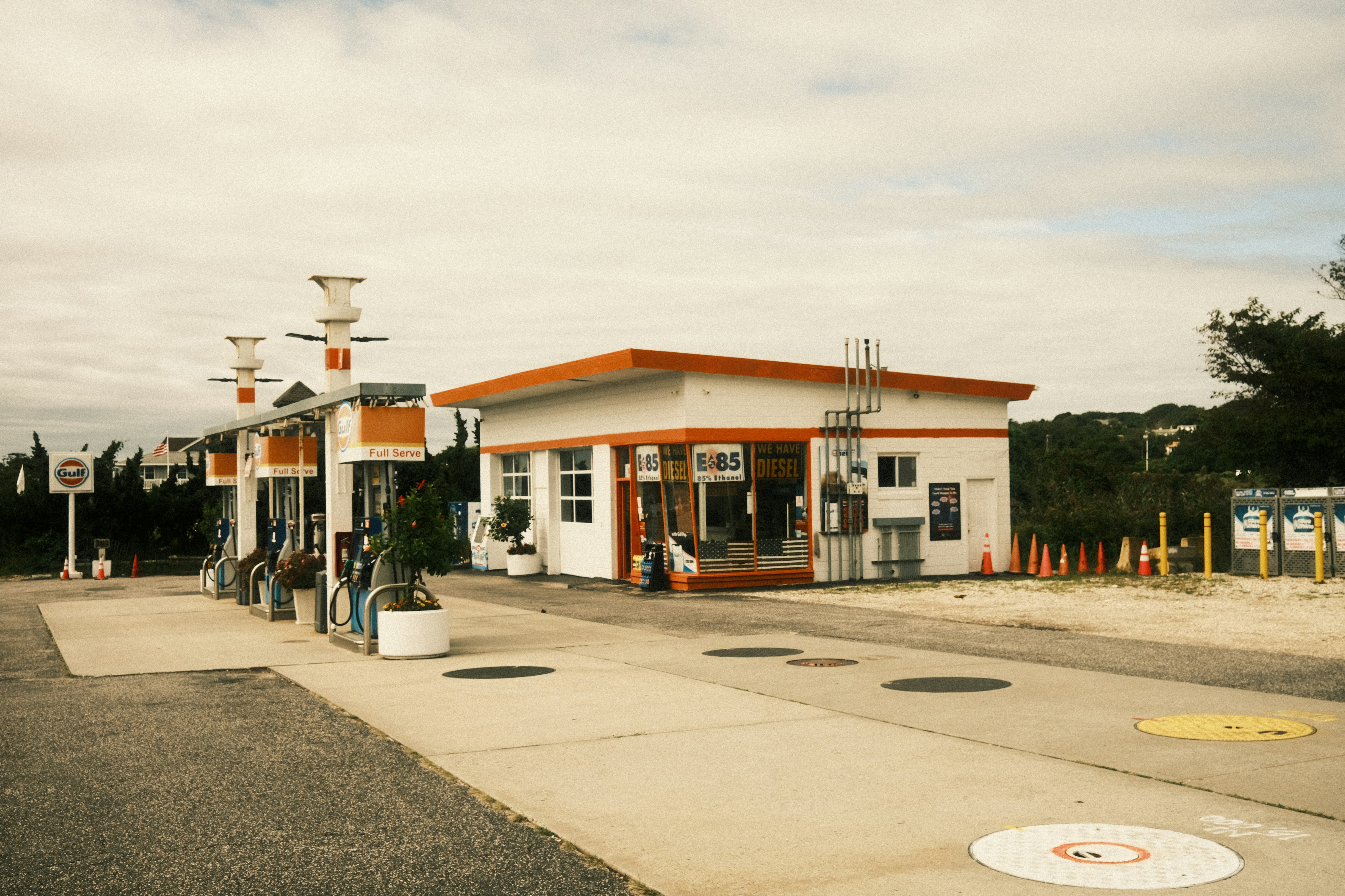 A retro-style gas station with orange accents sits along a quiet concrete forecourt, pumps on the left and a small building in the center; sky and distant landscape provide a calm backdrop.