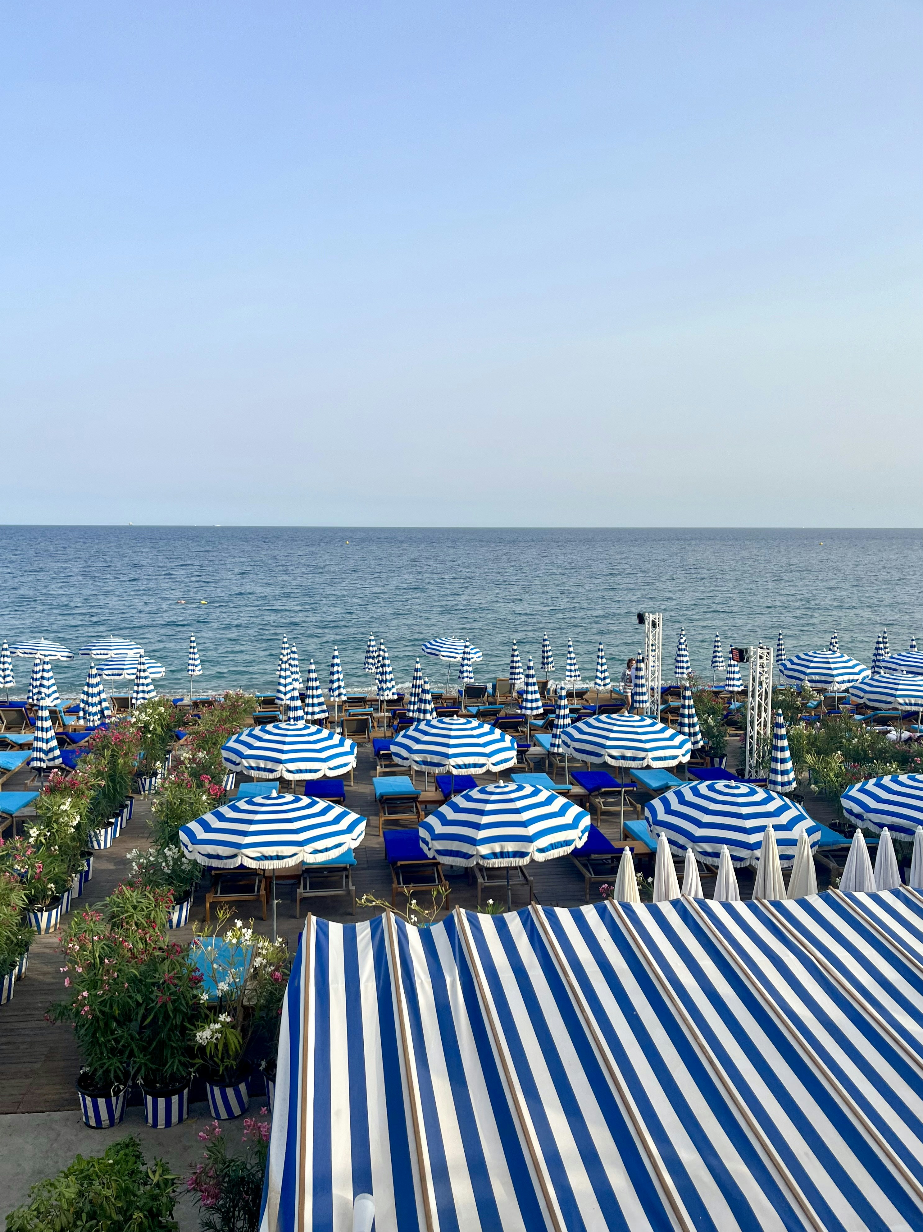 Rows of blue-and-white striped umbrellas line a beach club with a calm sea beyond, inviting a stroll toward the horizon. The scene is a photographed view of a seaside setup featuring repeated patterns and vibrant colors.