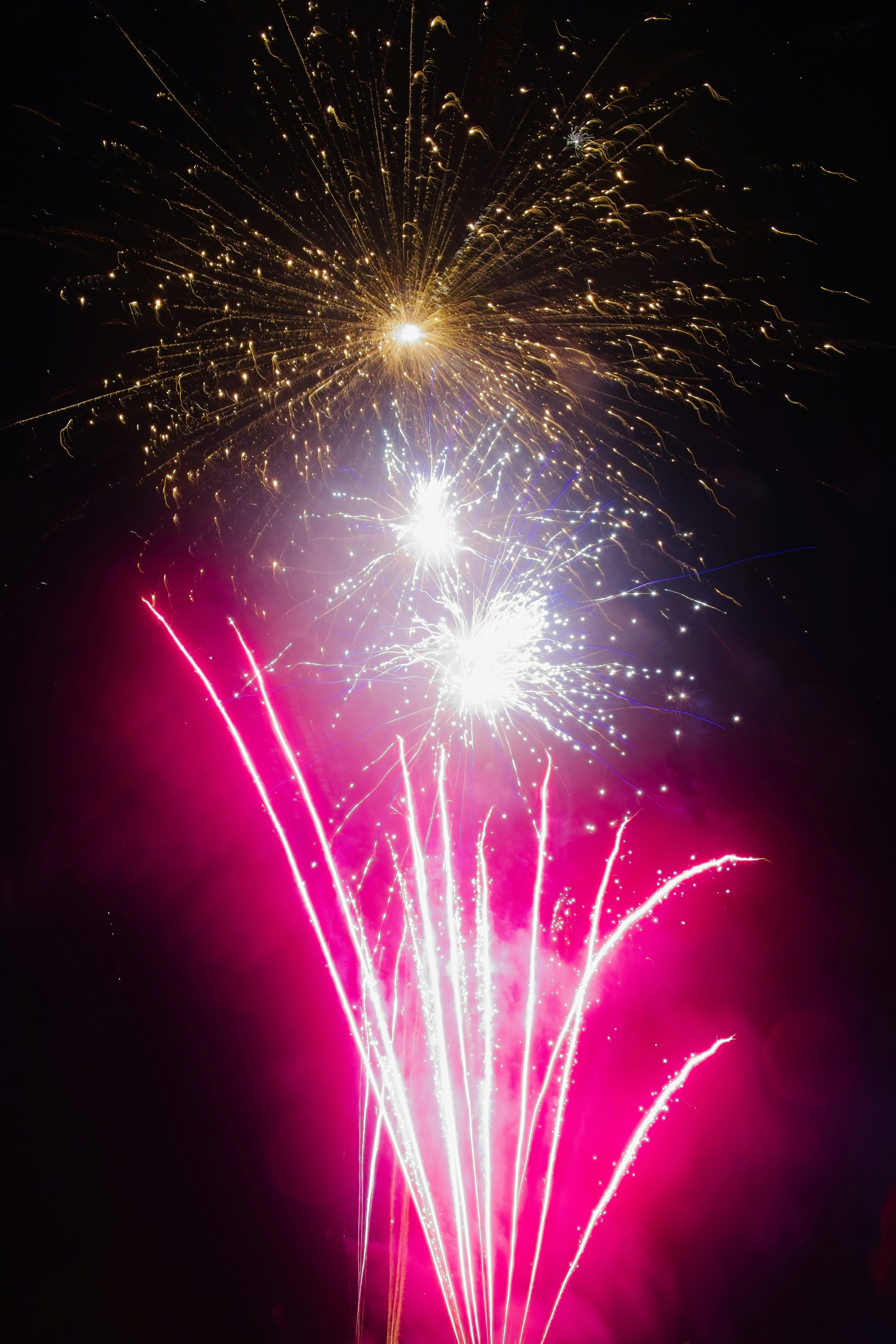 Colorful fireworks erupt against a dark sky, with neon pink arcs and white bursts creating a dramatic vertical display. This image captures a celebratory moment, emphasizing motion and light trails.