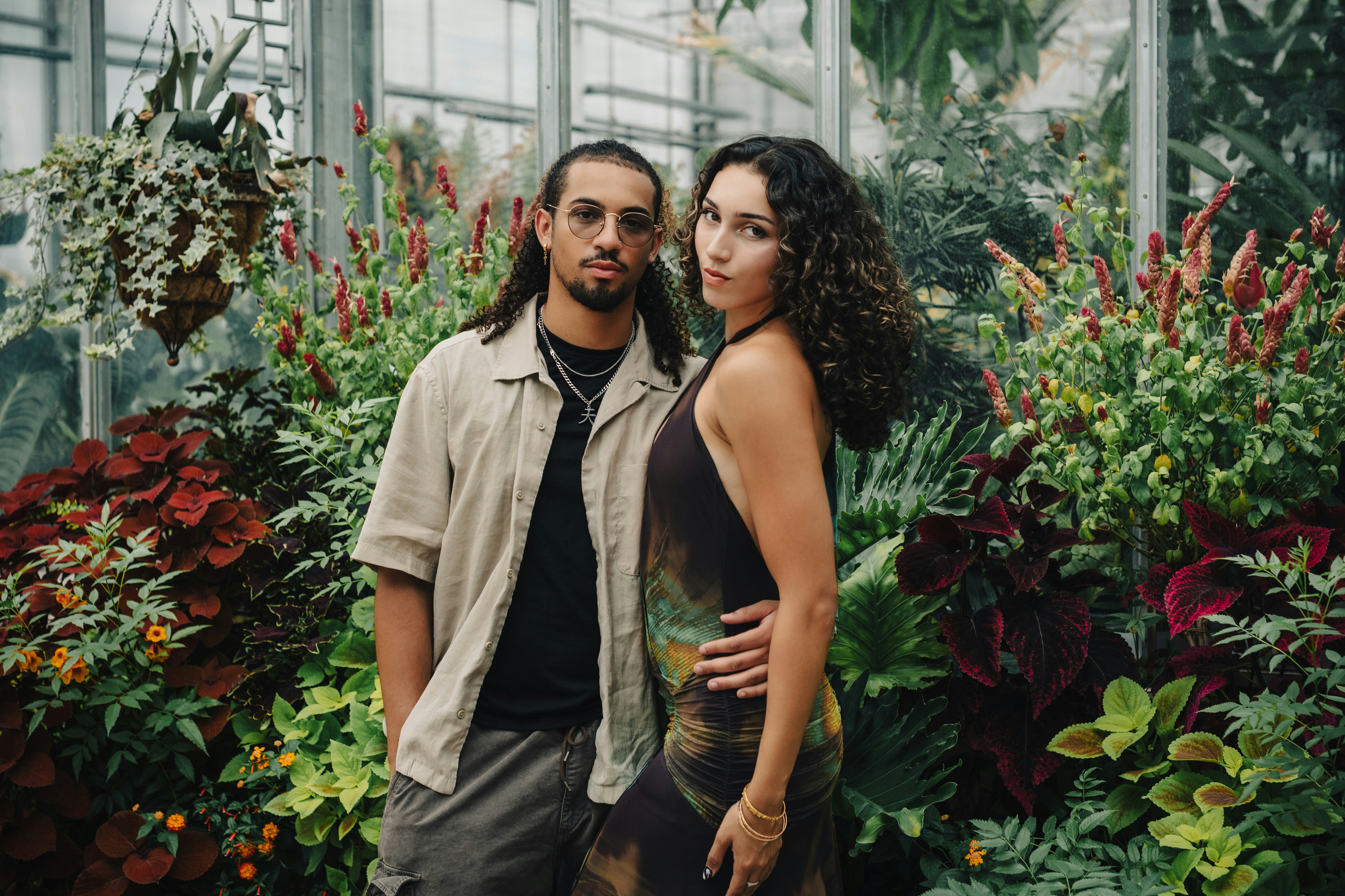 Two individuals pose closely among lush greenhouse foliage, showcasing fashion and connection in a vibrant botanical setting.