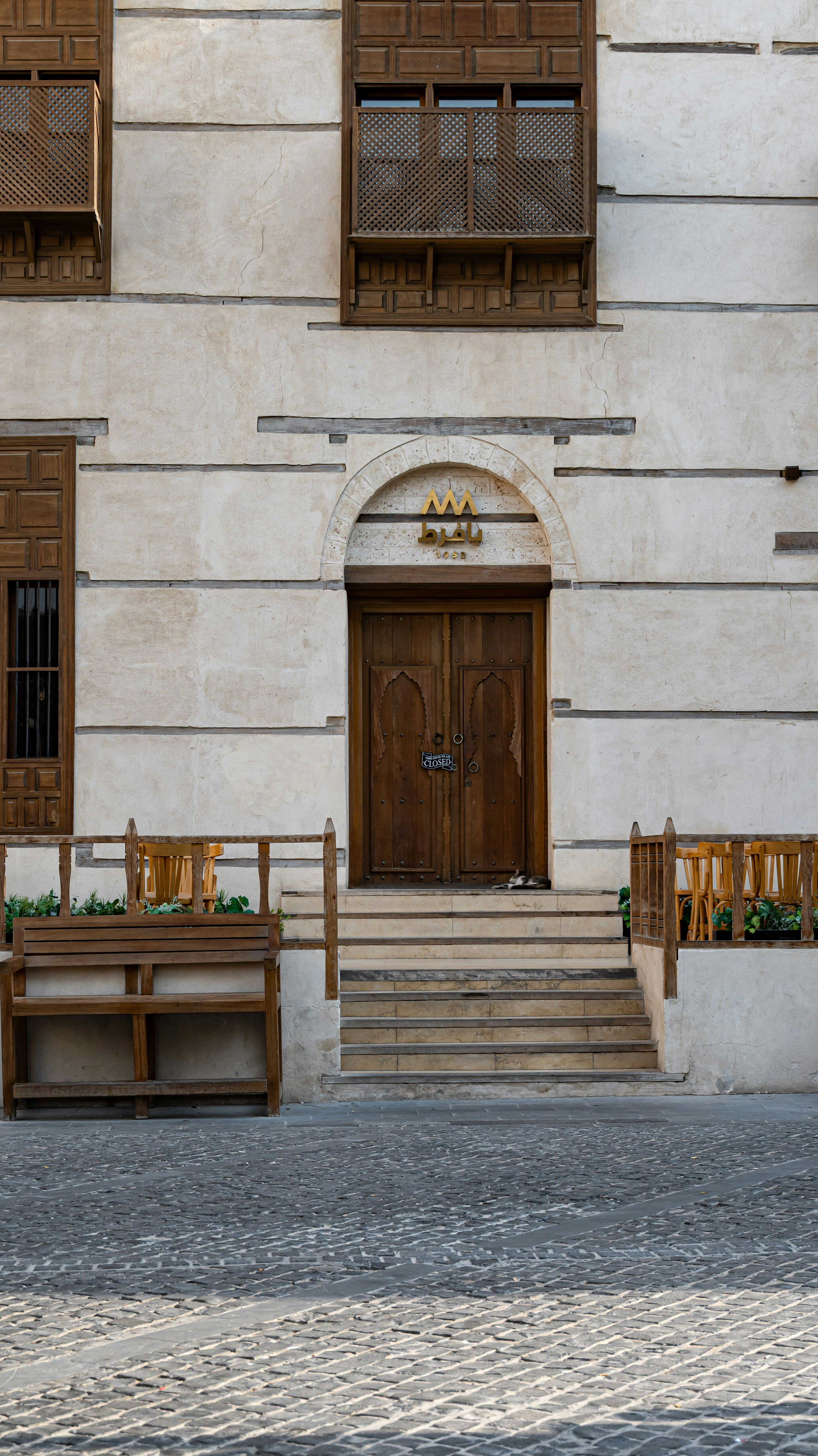 Stone facade with symmetrical wooden door and balconies, benches flanking a central staircase leading to an arched doorway; a calm, rustic street scene with muted tones.
