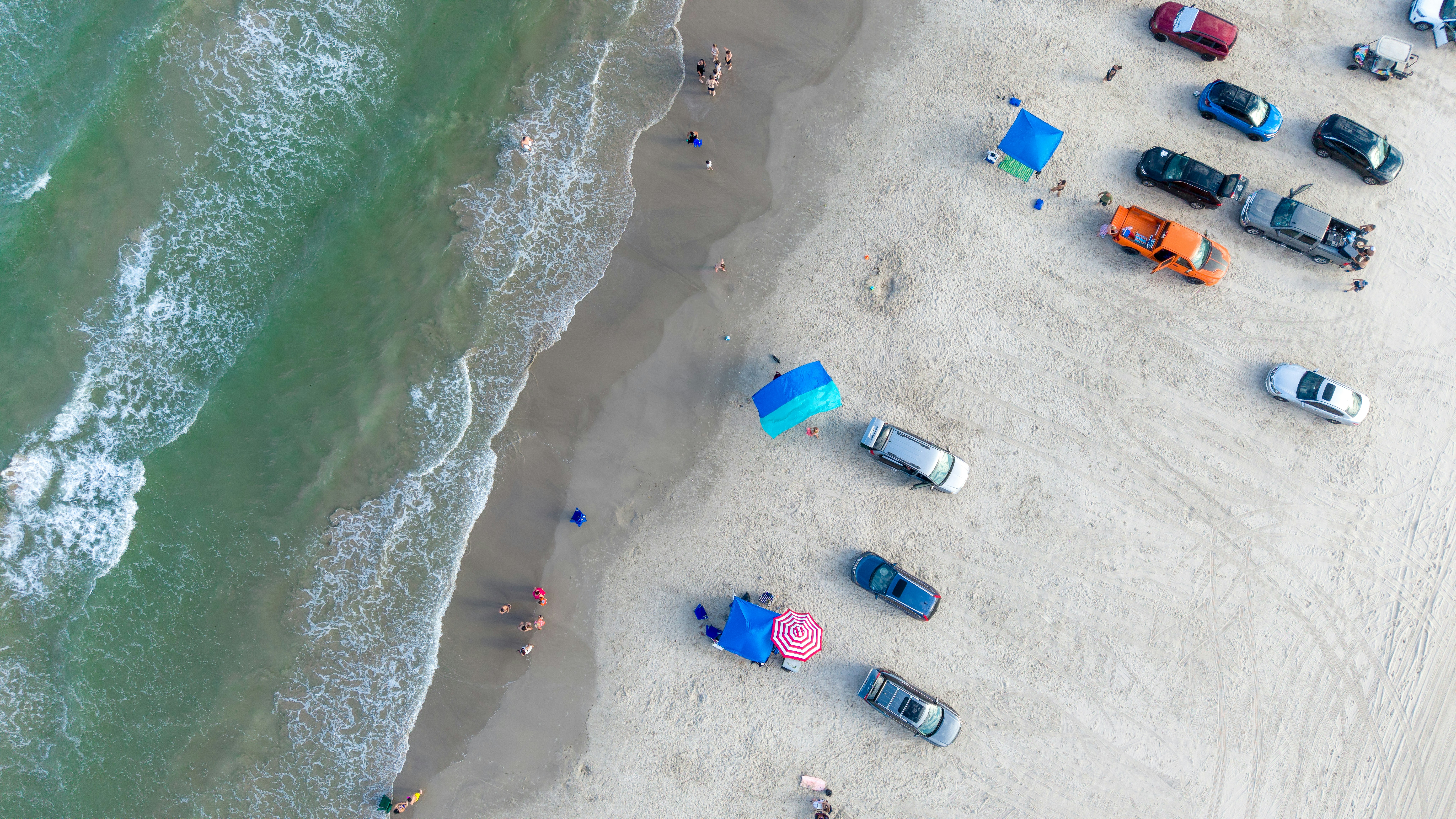 A drone shot shows a sandy beach with scattered cars, tents, and colorful umbrellas beside turquoise sea; vehicles line up on the sand, while beachgoers enjoy the water. Non-silhouette, natural daylight.