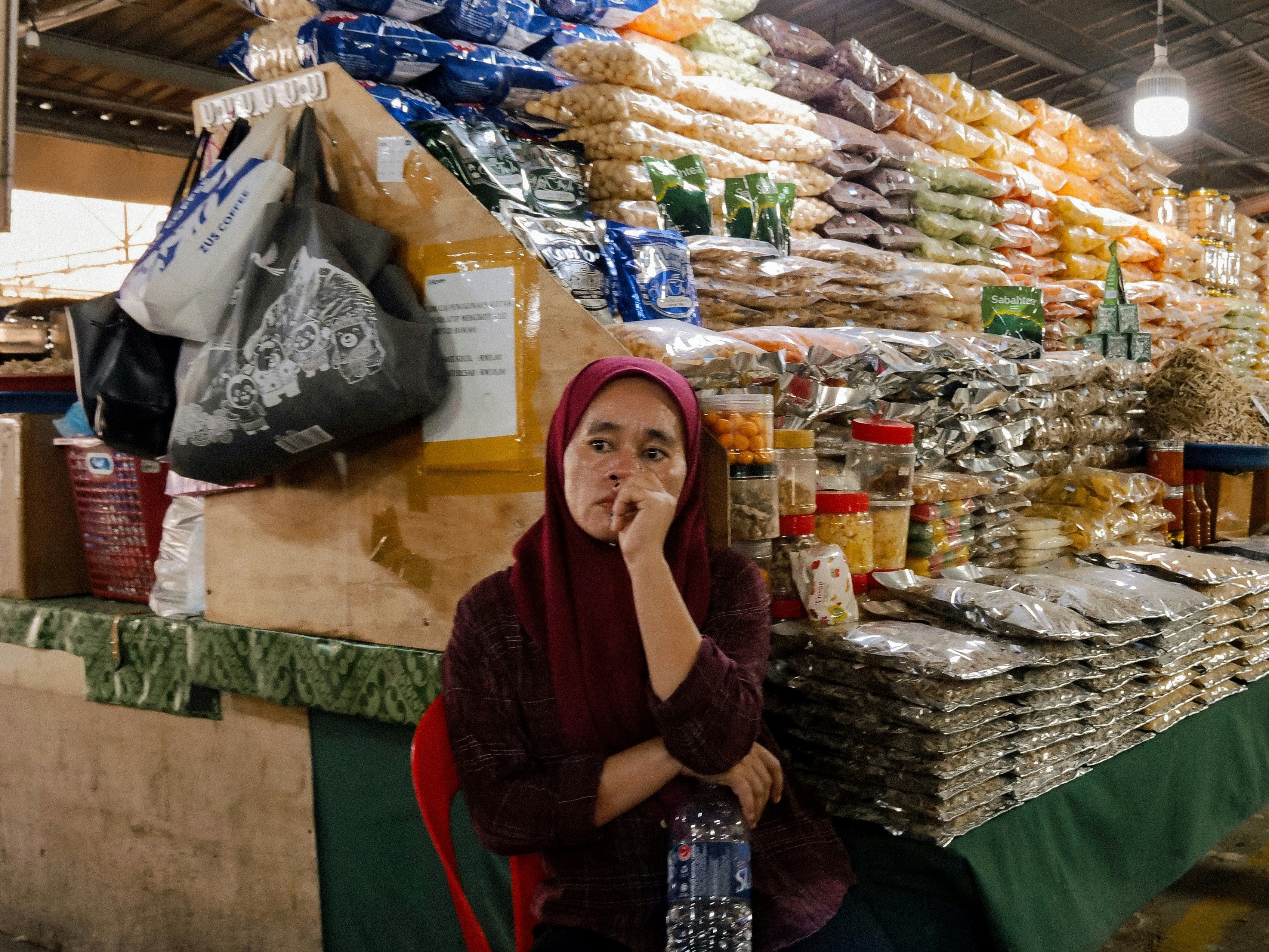 A woman with a red headscarf sits pensively at a vendor stall stacked with colorful goods in a busy market; the scene emphasizes human stillness amid abundance.