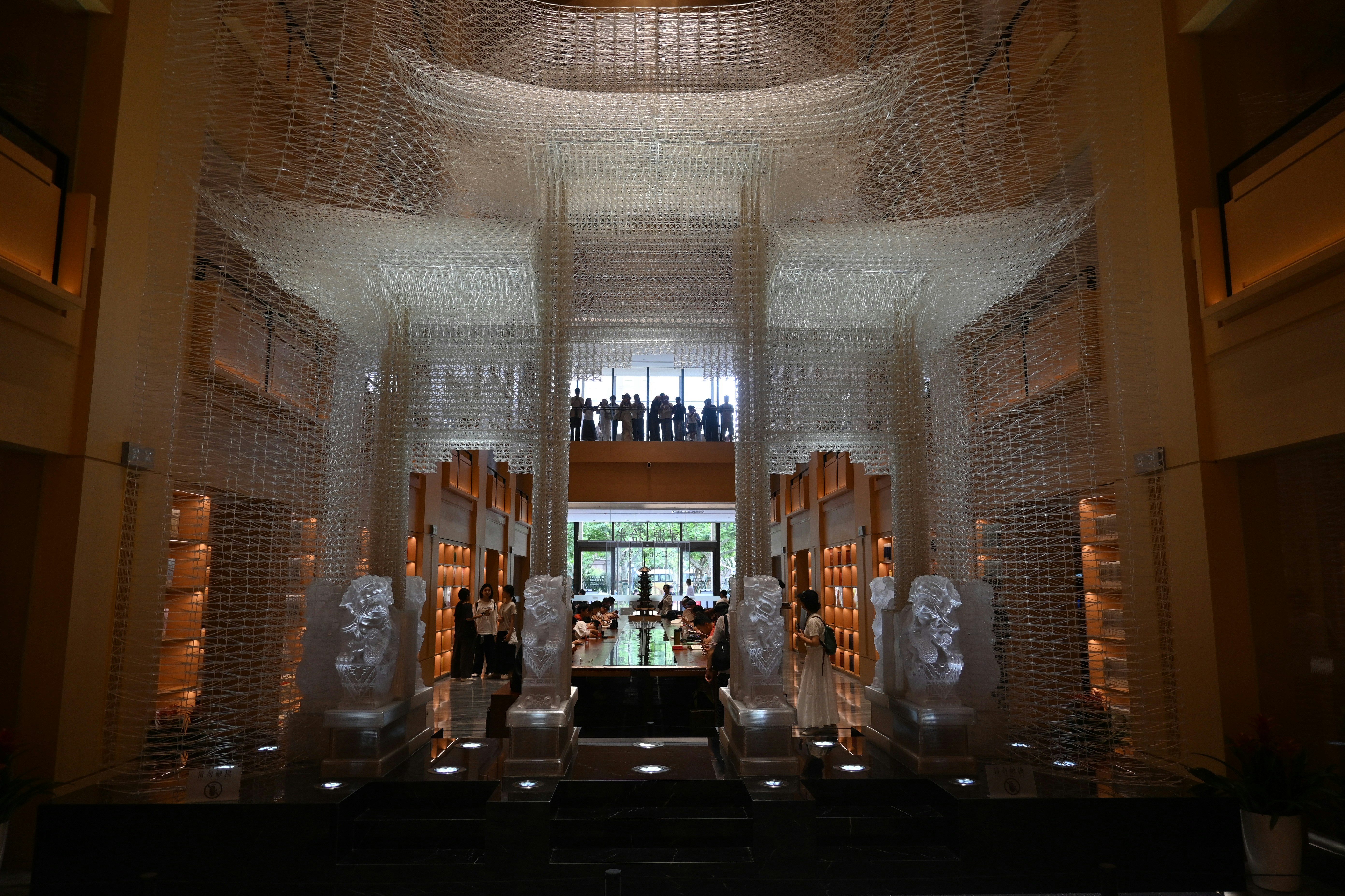 Wide interior with mirrored walls and statues lining a central aisle, viewers gathered on a balcony above, creating a sense of depth and reverence.