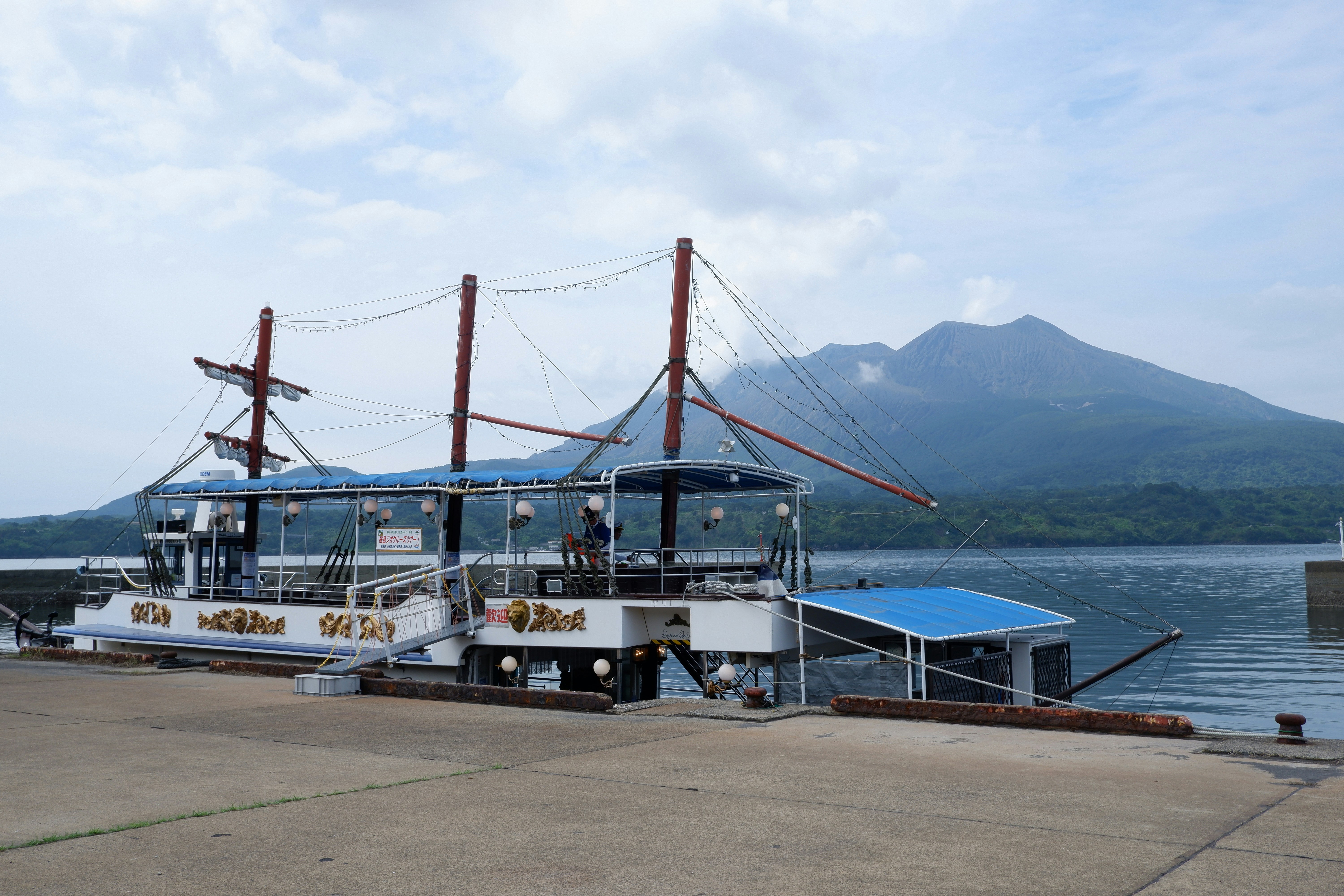 A docked boat with a blue canopy rests by a calm shore, with distant mountains under a partly cloudy sky.