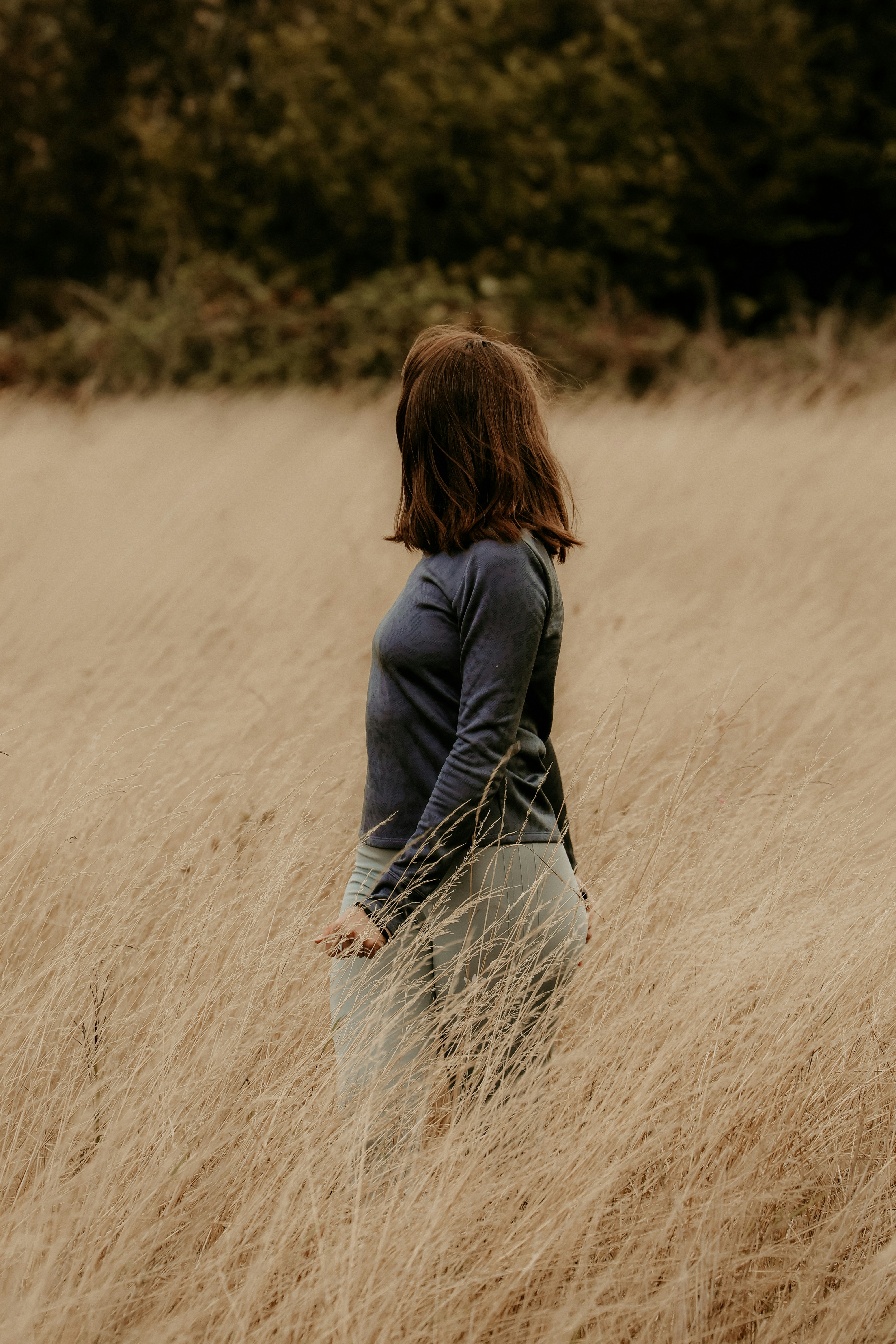 A woman stands in a sunlit field of tall, golden grass, facing to the left with soft, natural tones. The scene emphasizes solitude and the gentle textures of the landscape; the image is a candid, photographic depiction rather than an illustration, logo, or painting.