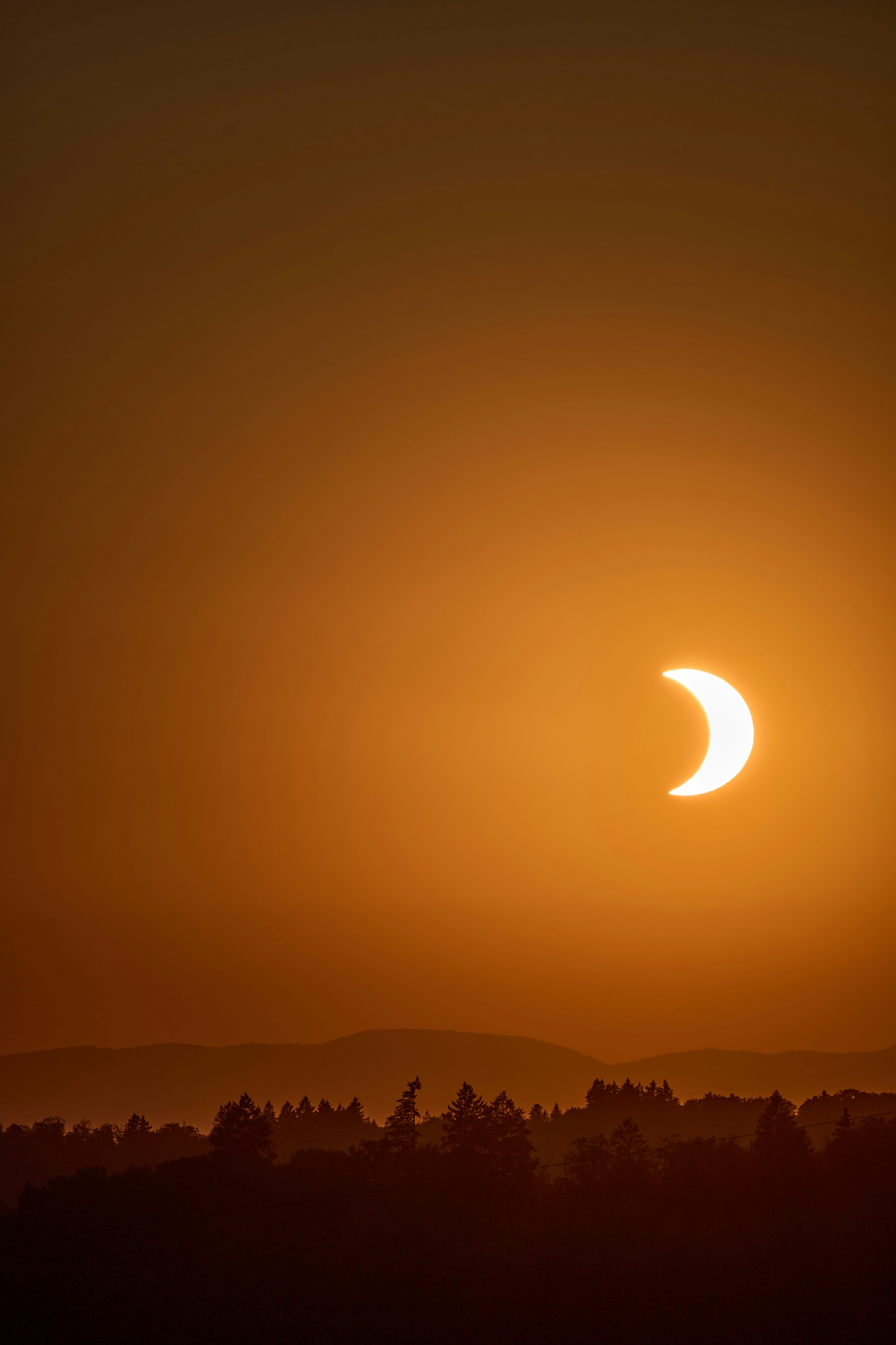 Deep orange horizon with a thin crescent sun above a silhouetted forest and rolling hills.