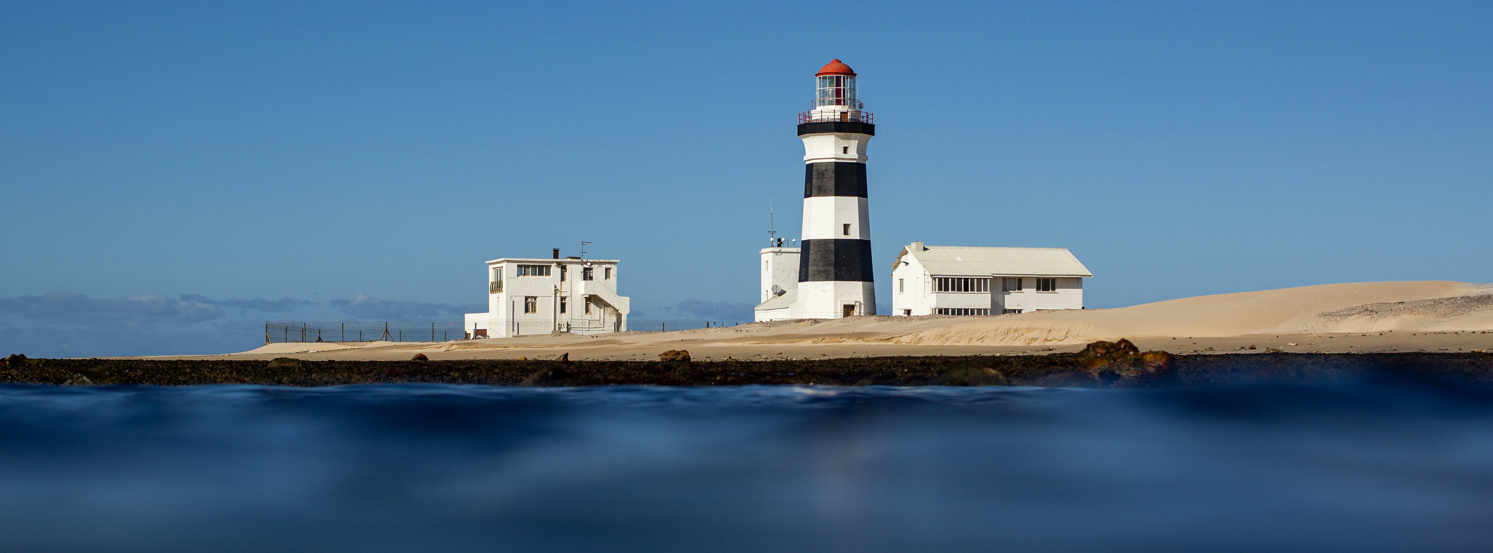 Lighthouse on a sandy shore with adjacent white buildings, set against a calm sea under a clear sky.