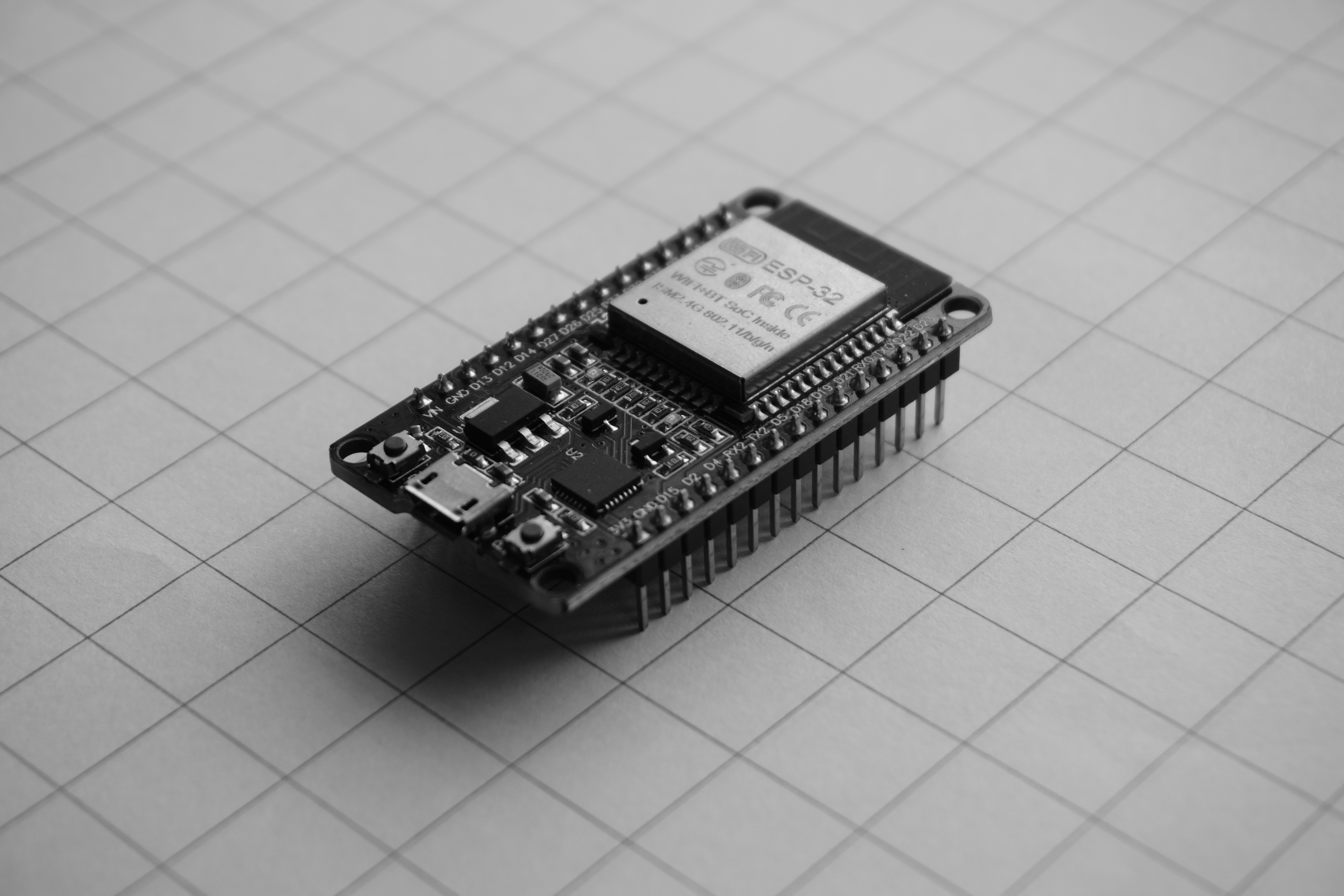 Small ESP32/ESP8266-like development board resting on a grid surface, highlighting its module layout and edge connectors.