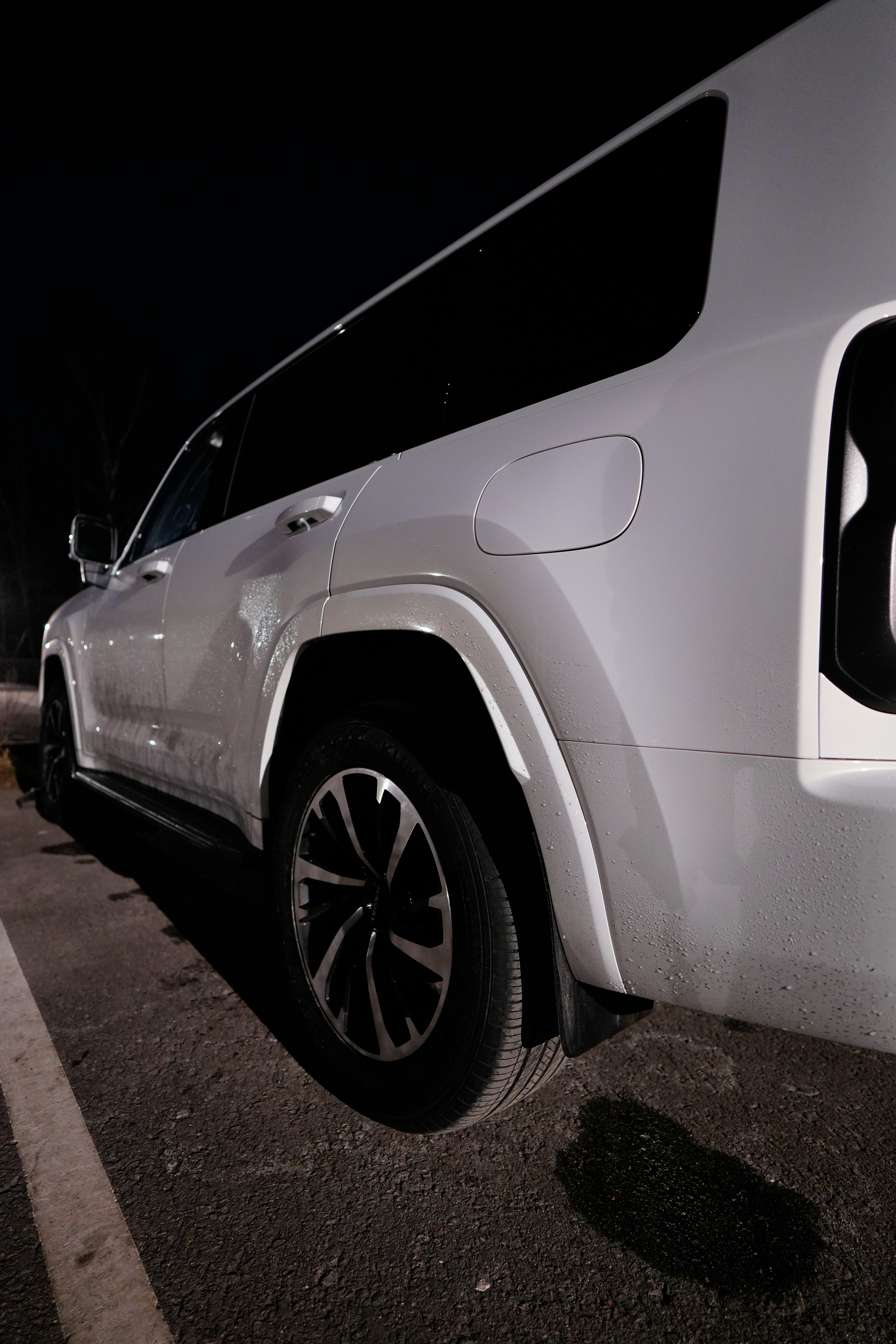 Low-light shot of a white SUV parked on a dark street, showcasing a close-up of the rear wheel and side panel with water droplets on the body.