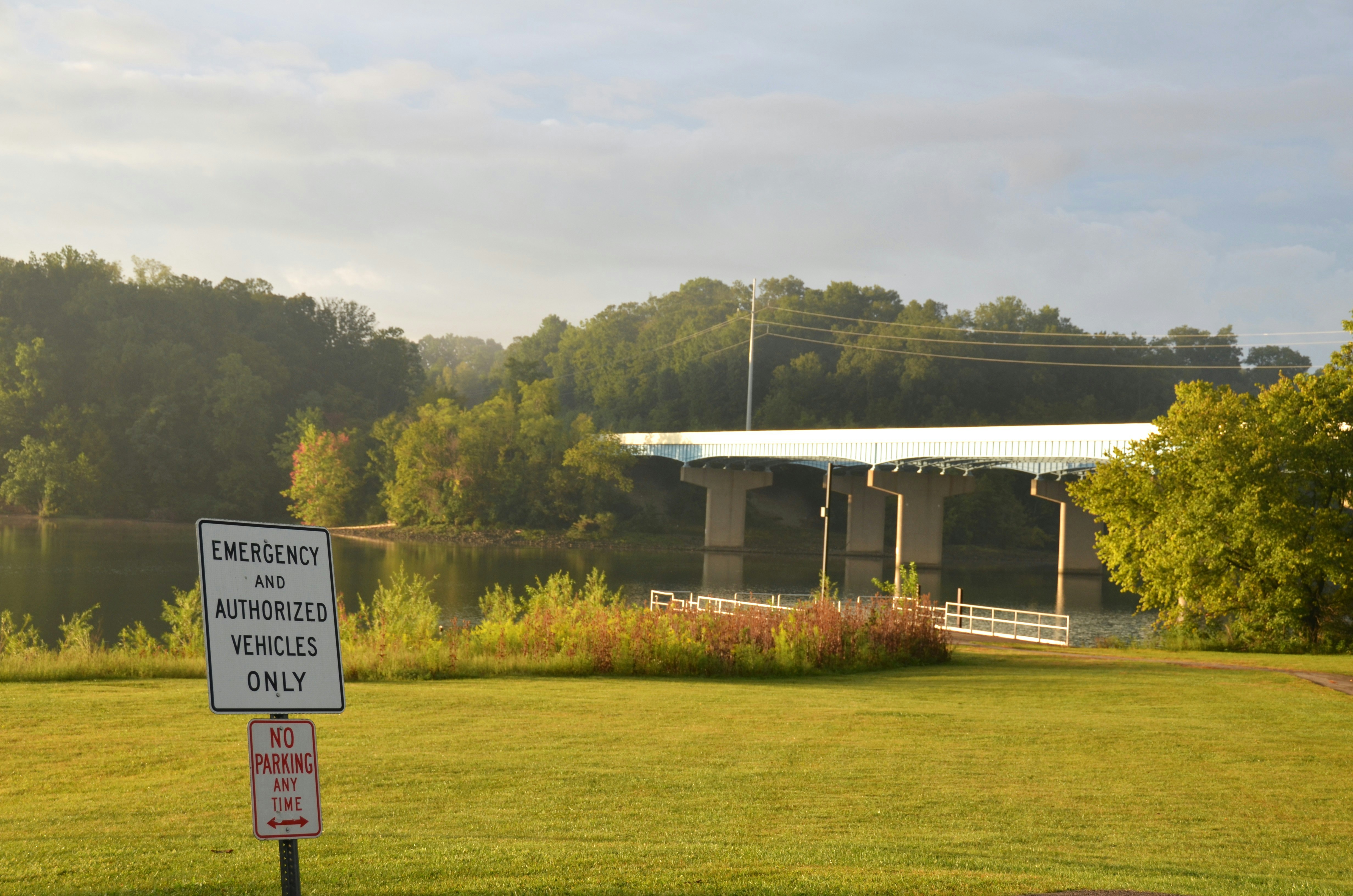 A tranquil riverside scene with a white bridge spanning a calm waterway and a manicured grassy foreground; an emergency vehicle sign sits near the lower left, giving a subtle urban context to the landscape.