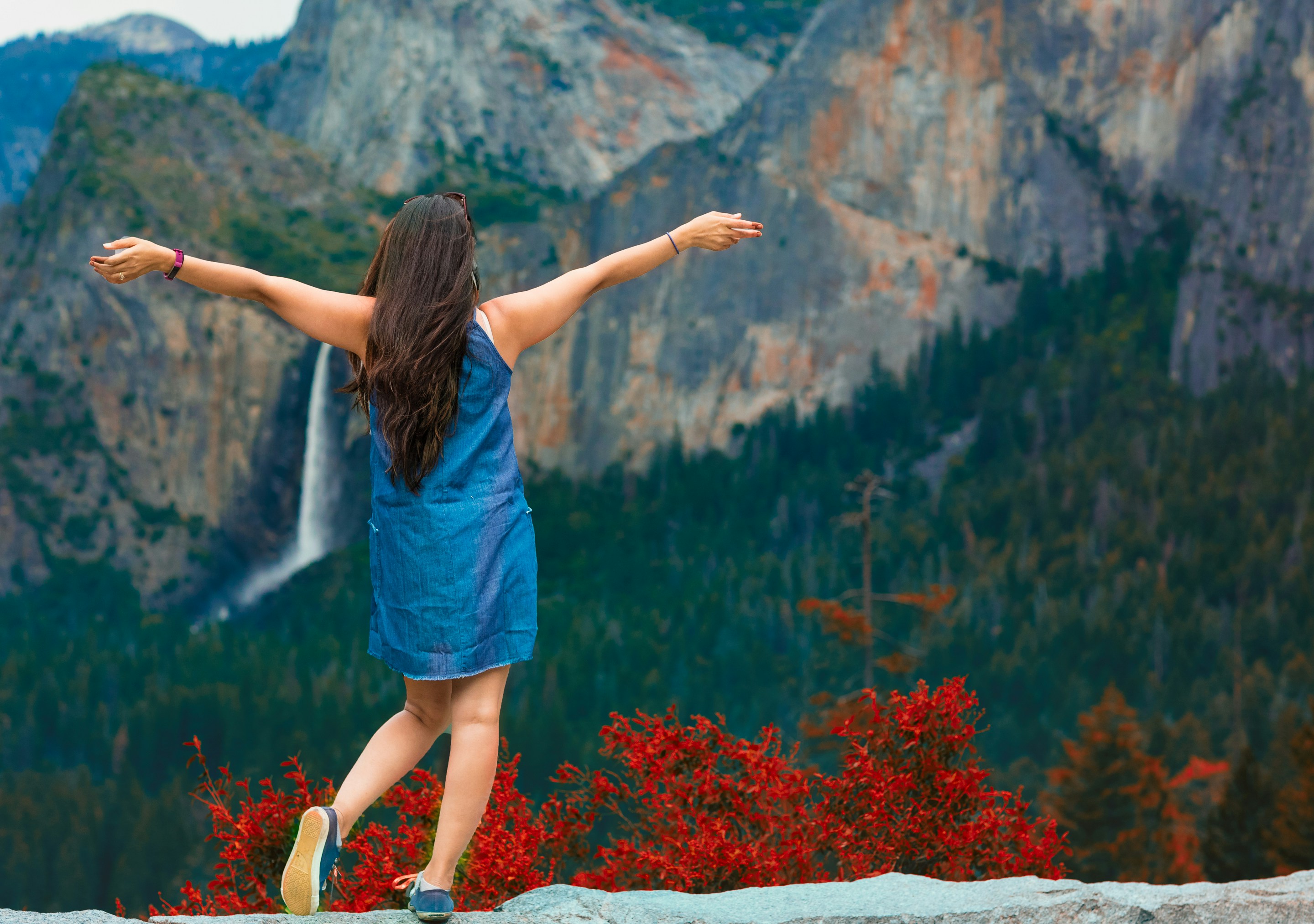 Young woman in a blue dress stands with arms outstretched on a rocky overlook, facing a waterfall and mountains amid autumn colors. This is a photographic capture of a person in a scenic landscape.