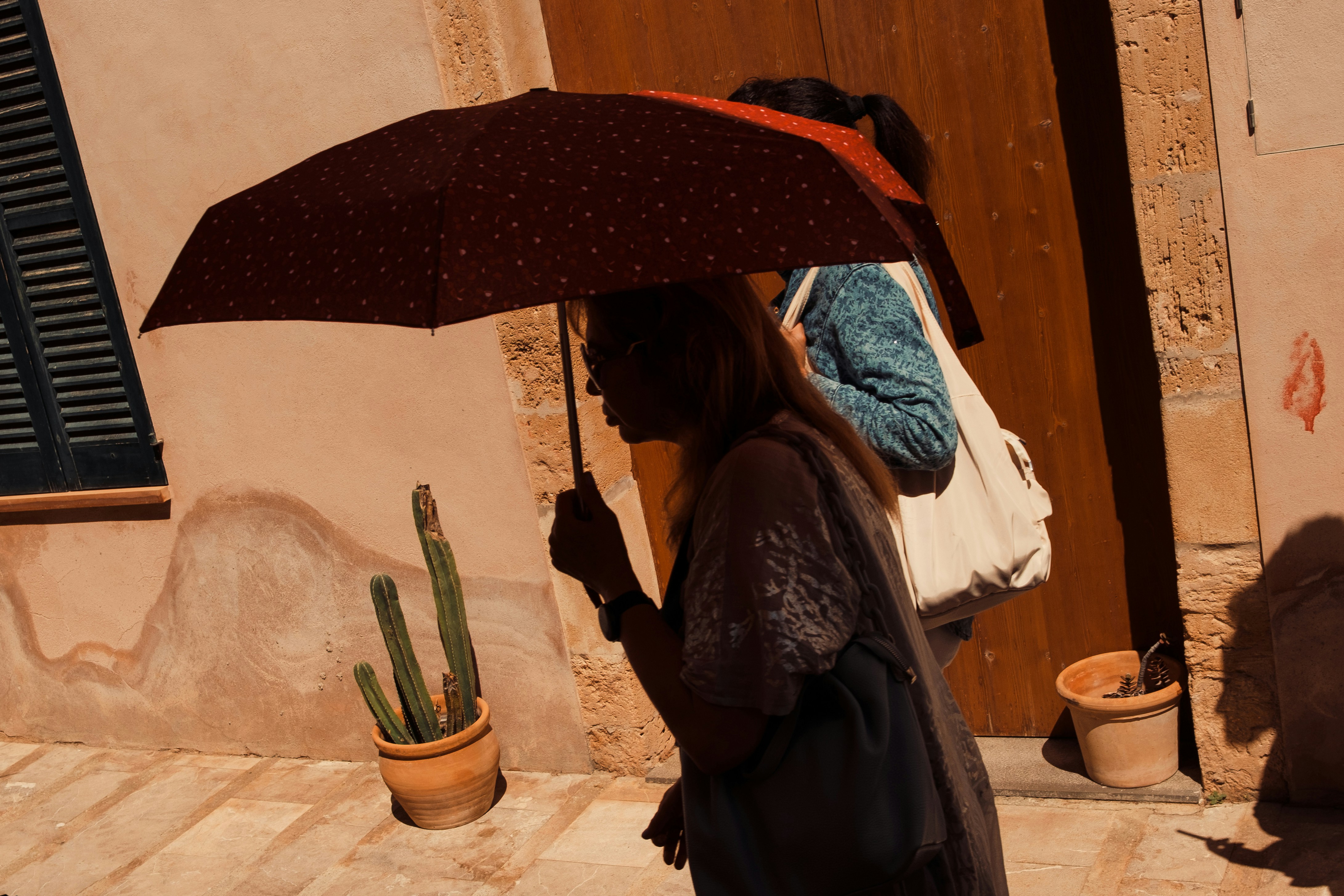 A warm, cinematic street photograph of two women walking past sandstone walls in the old town of Alcúdia, Mallorca, shielded by a patterned red sun umbrella in bright Mediterranean light.
