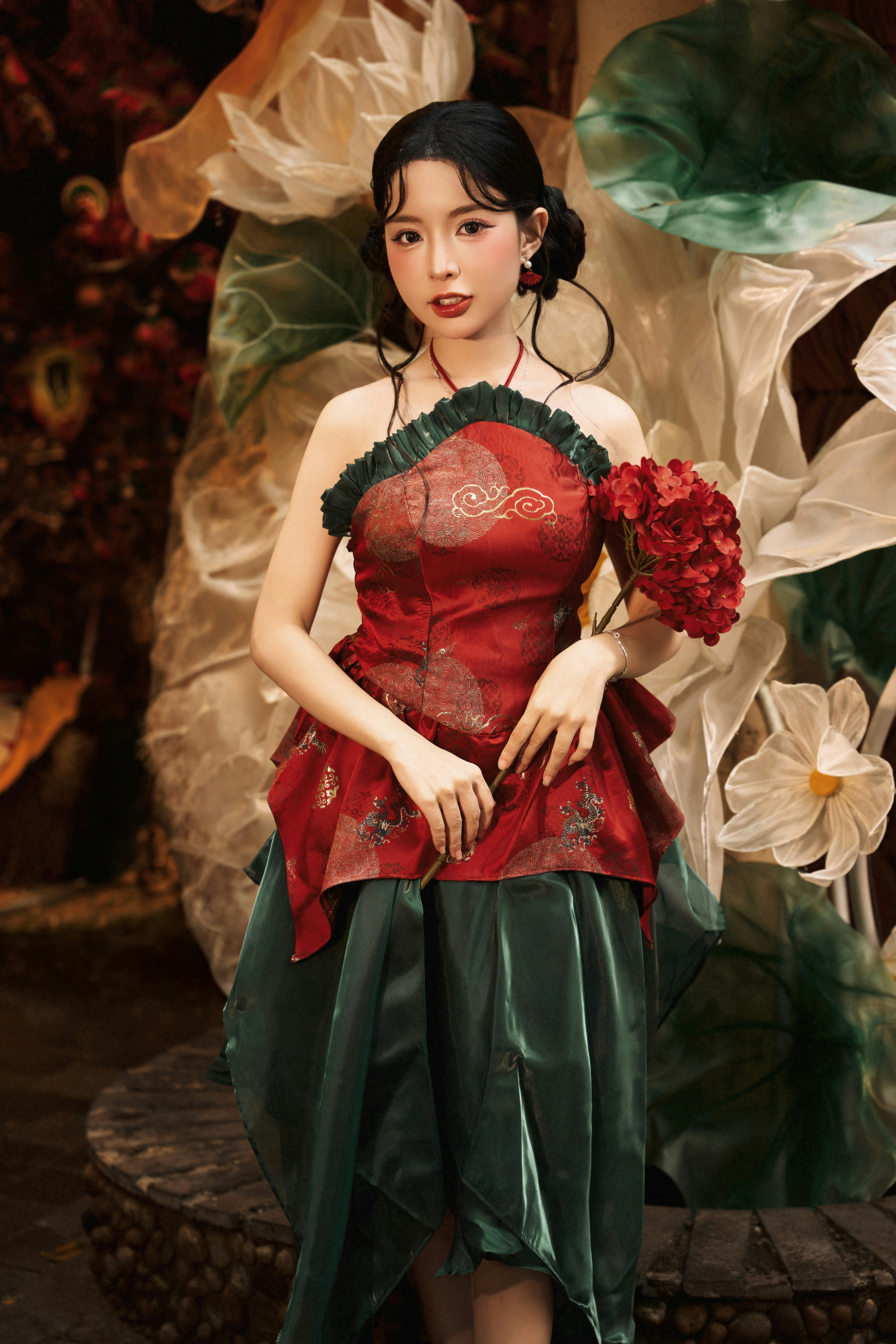A poised woman in a red and green gown stands among oversized floral props, holding a red bouquet. The scene emphasizes ornate costume and soft, theatrical lighting as a portrait of performance style.
