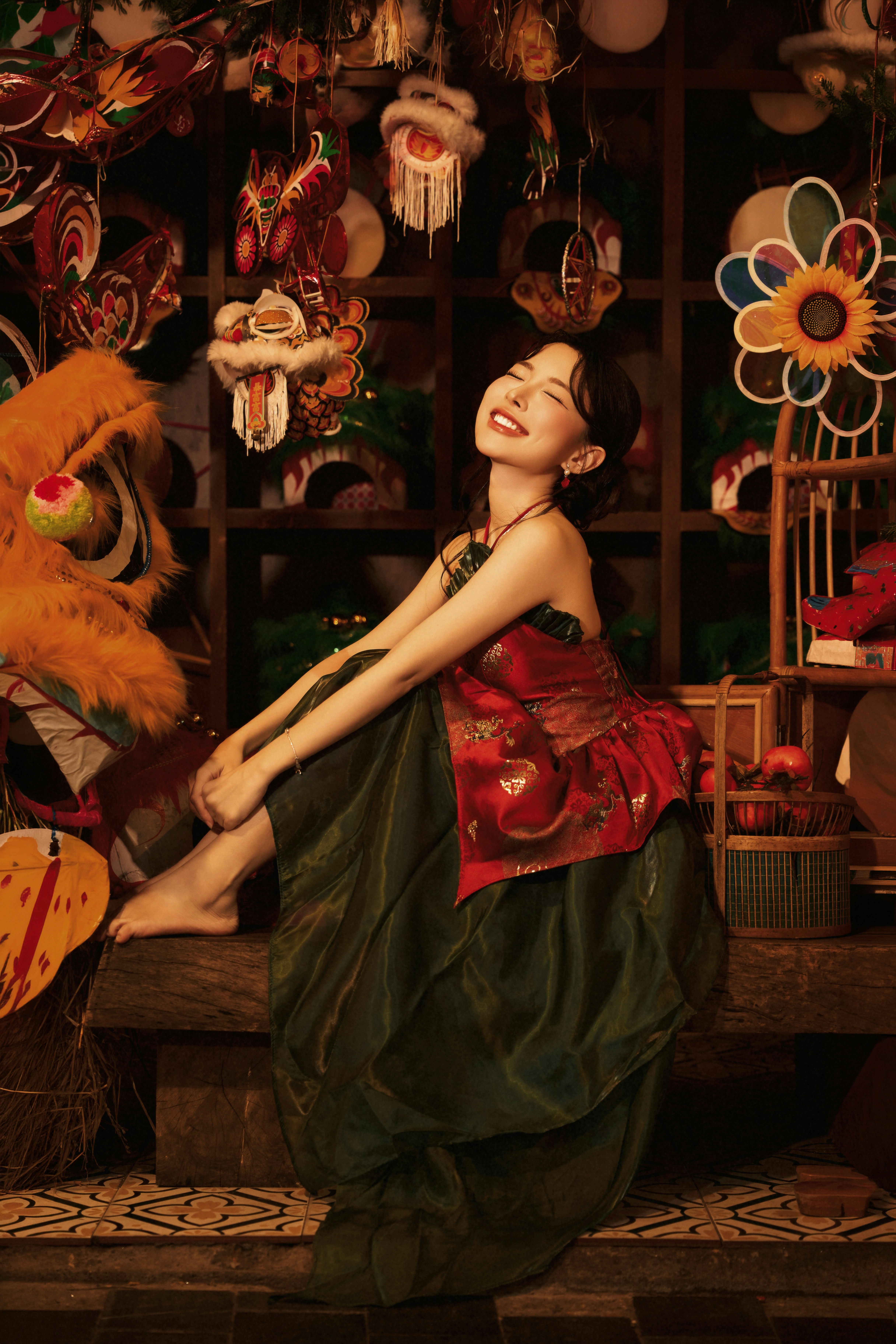 A smiling woman in a lush red and green gown sits on a wooden platform amid festive decorations and puppets. The scene exudes warm, intimate holiday charm with soft lighting and rich textures.