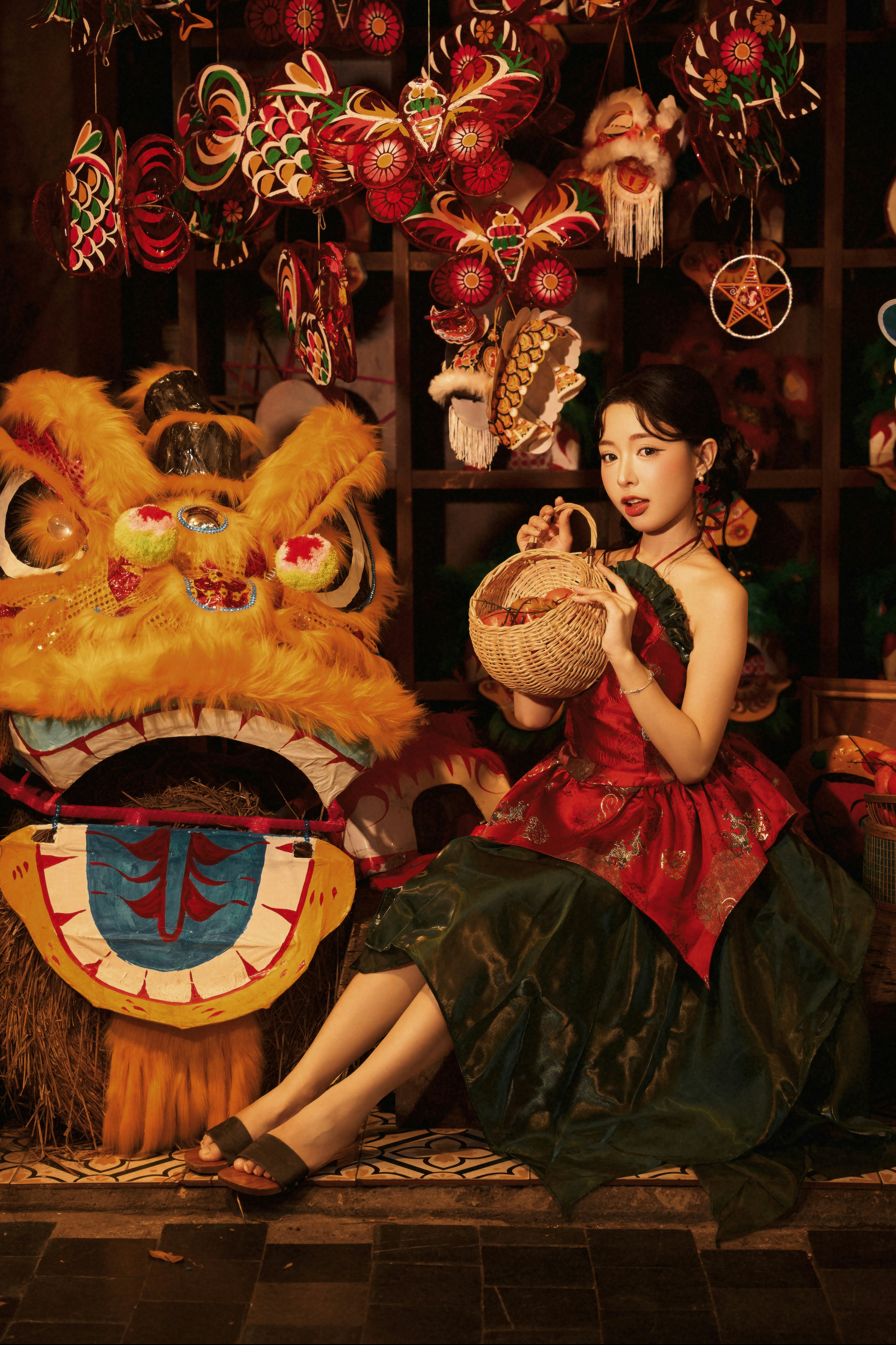 Woman in a red-and-green dress sits beside a vibrant yellow dragon figure, holding a woven basket amid hanging festival decorations in warm lighting.