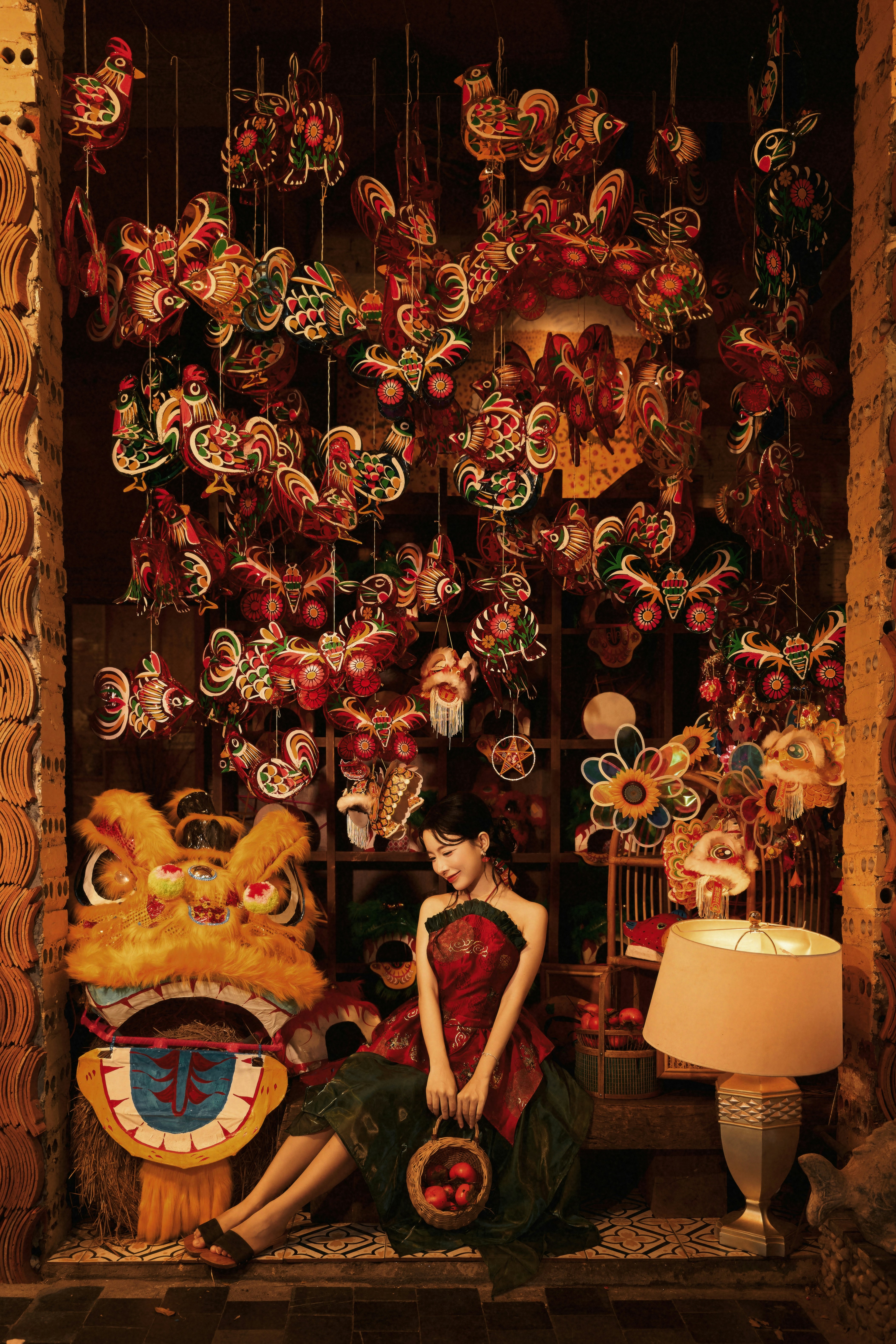 A gracefully dressed woman sits among colorful hanging kites and decorative props in a warmly lit studio-like space; a lion head and retro lamp frame the scene, suggesting a staged cultural tableau.