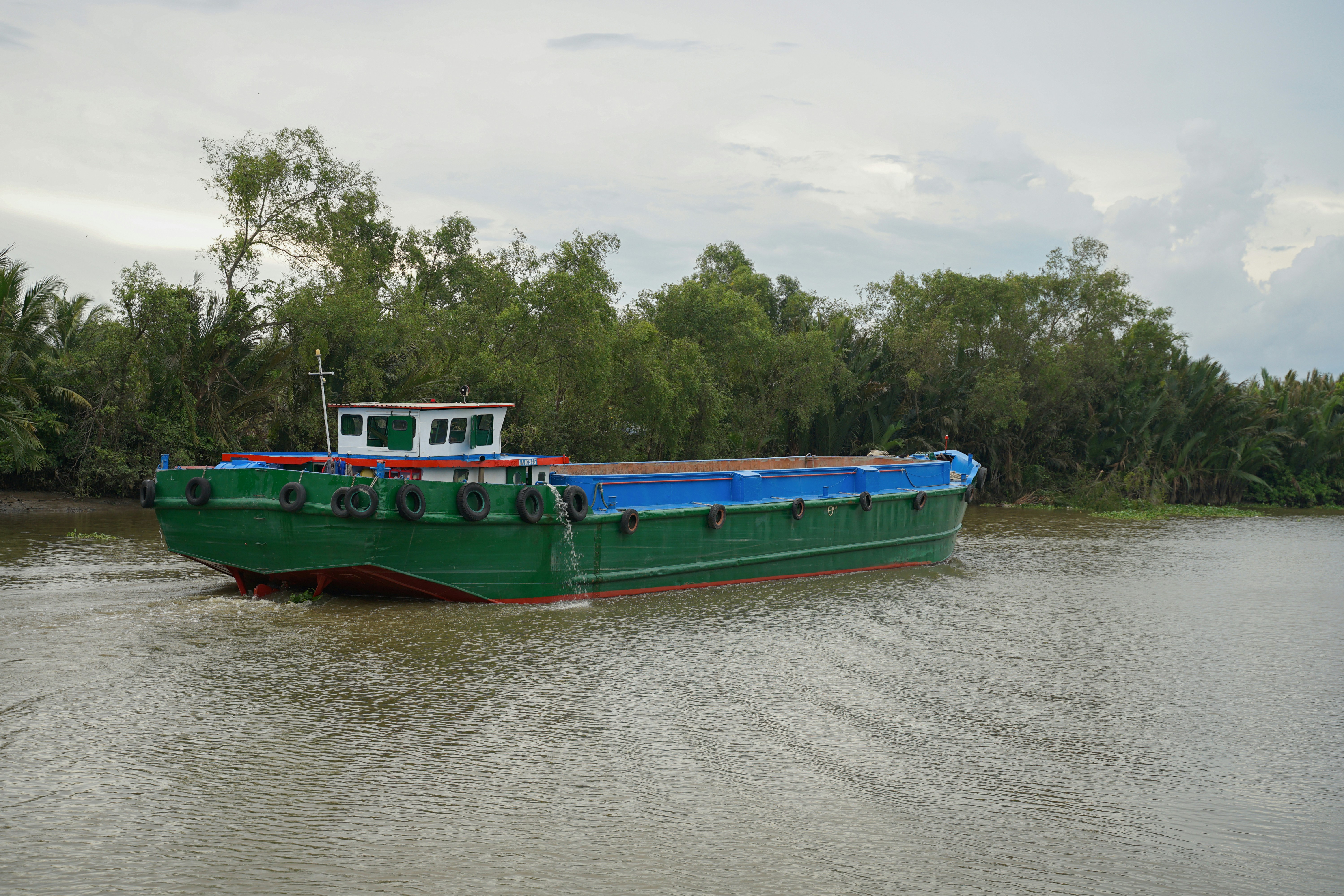 A green and blue cargo barge drifts on a wide river with dense trees in the background; the vessel occupies the left-half of the frame, conveying a tranquil, rural waterway scene.