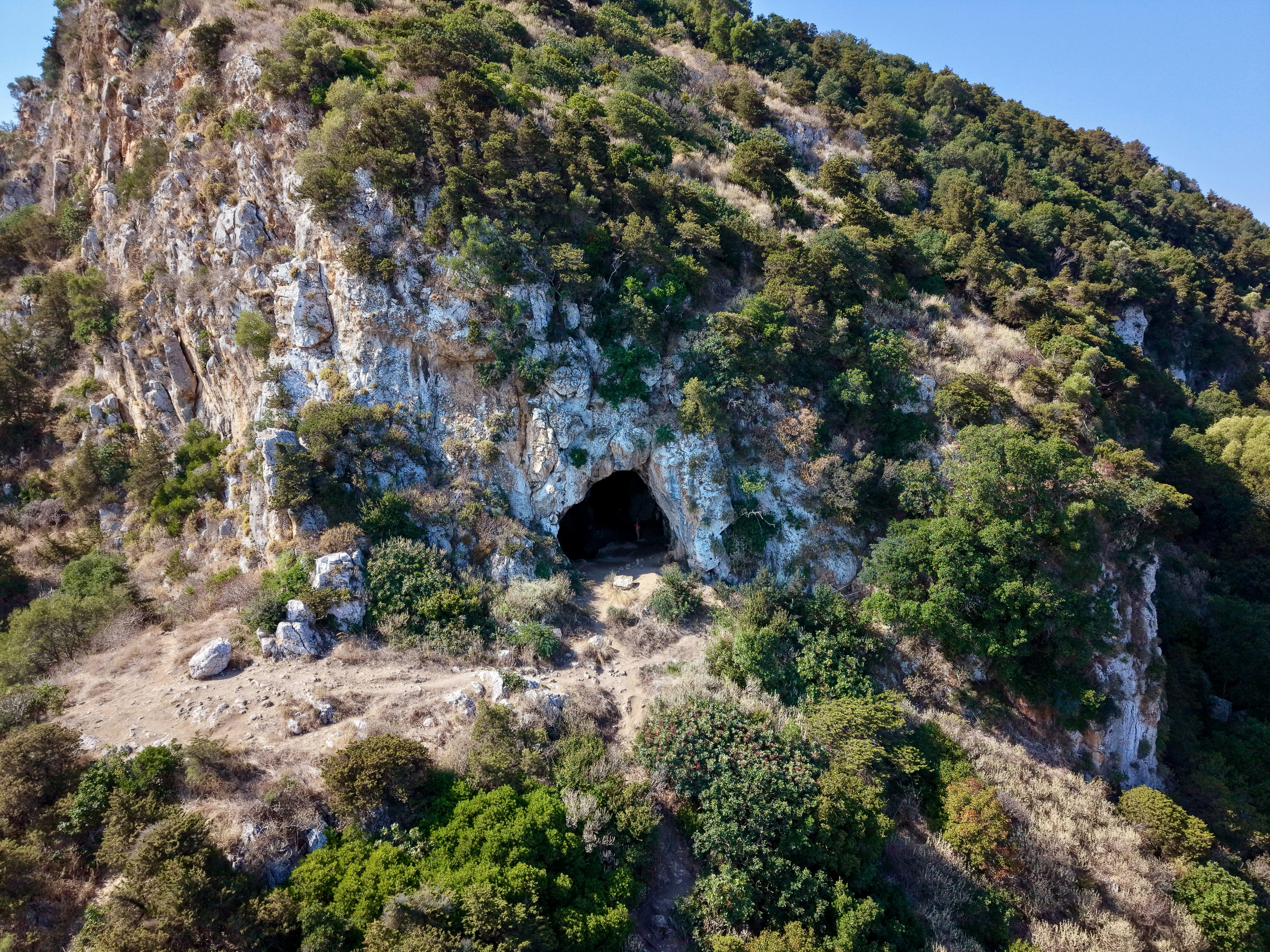 A dark cave entrance in a rocky cliffside covered with green trees and shrubs