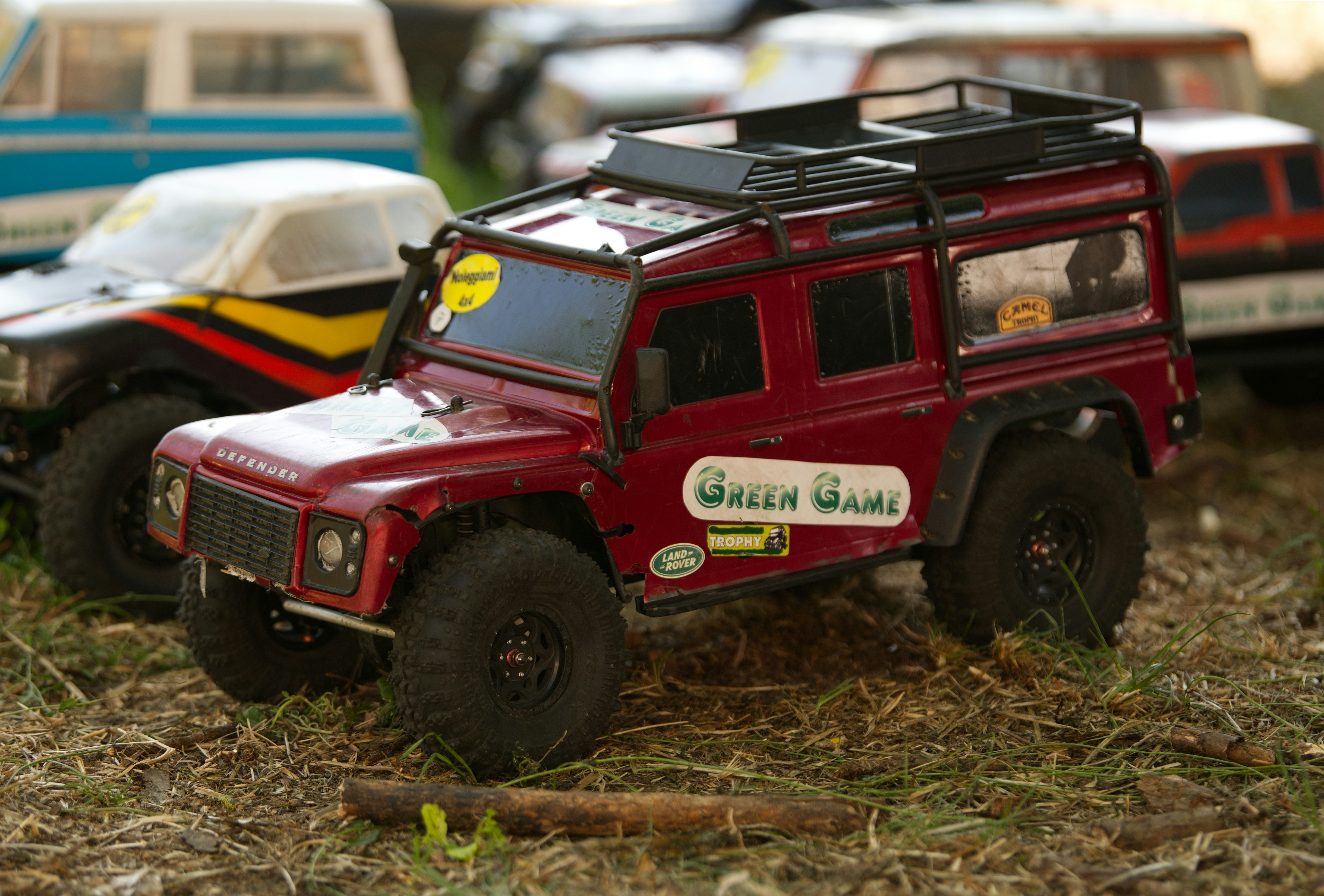 Compact red toy off-road SUV with stickers and roof rack sits on dirt and grass, surrounded by other model vehicles in a toy landscape.