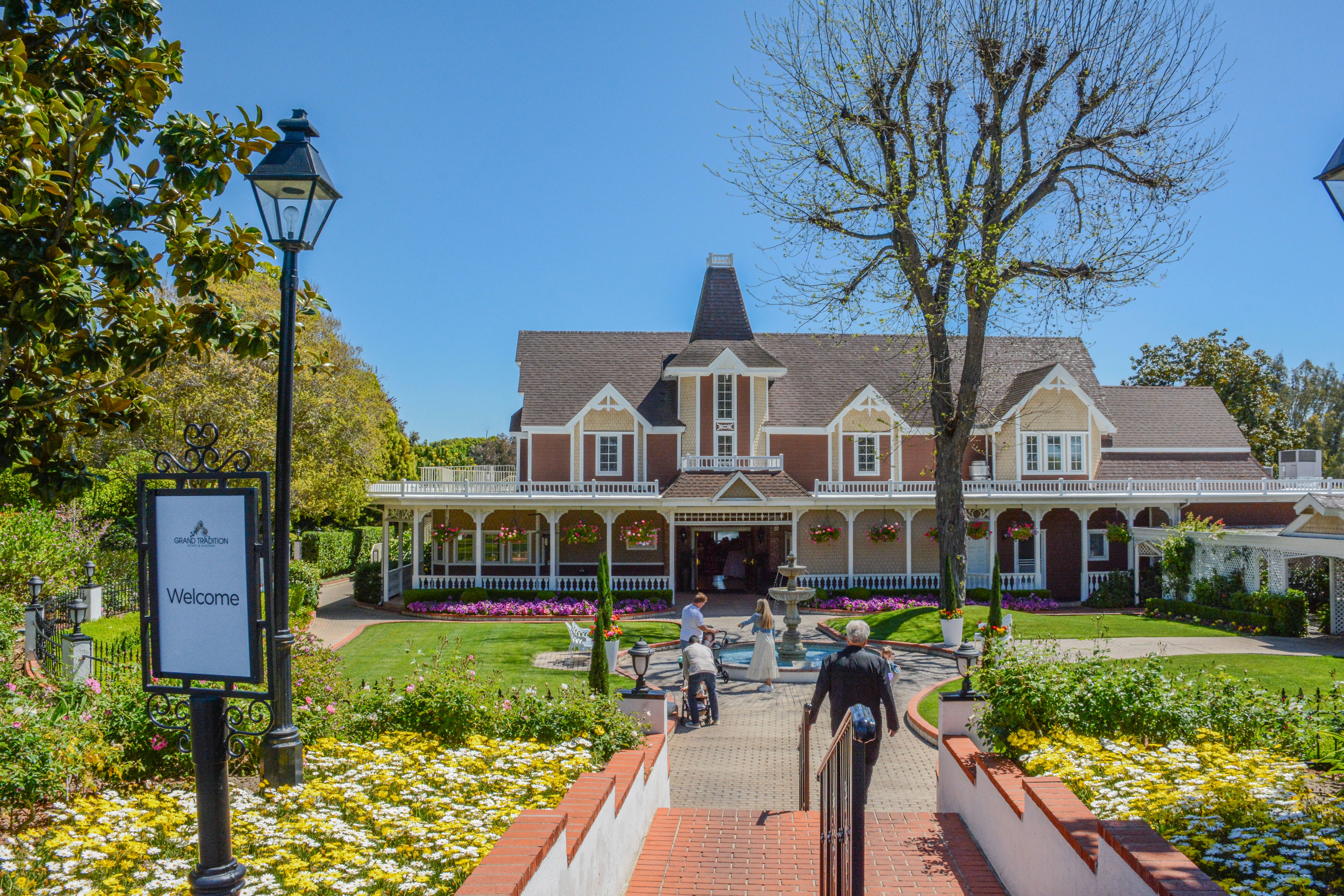 People walking through a garden toward a large house with a fountain