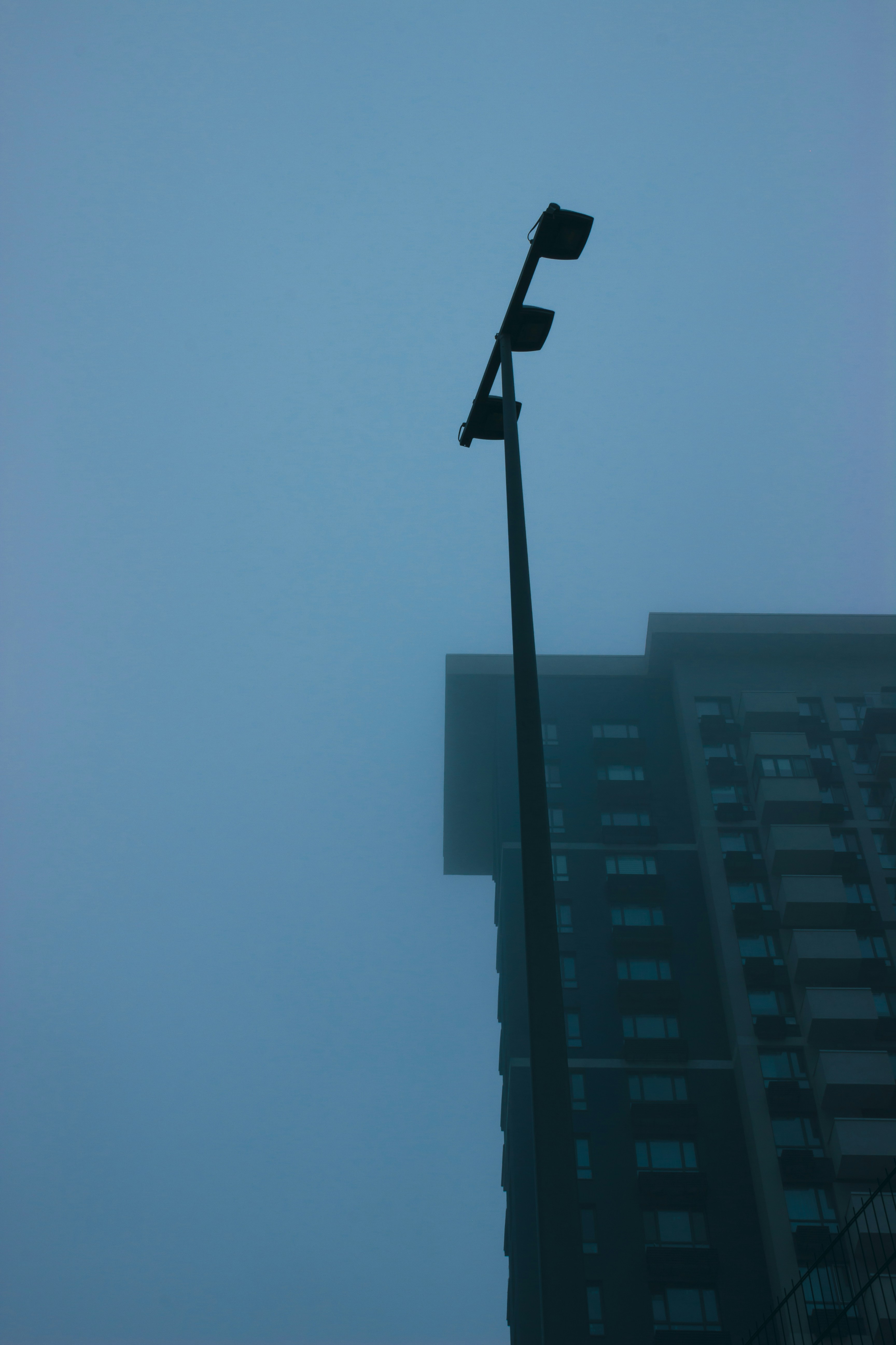 Blue-tinted cityscape scene with a tall street lamp rising beside a modern building silhouette in heavy fog. Abstract urban architecture and minimal lighting create a moody, atmospheric composition.