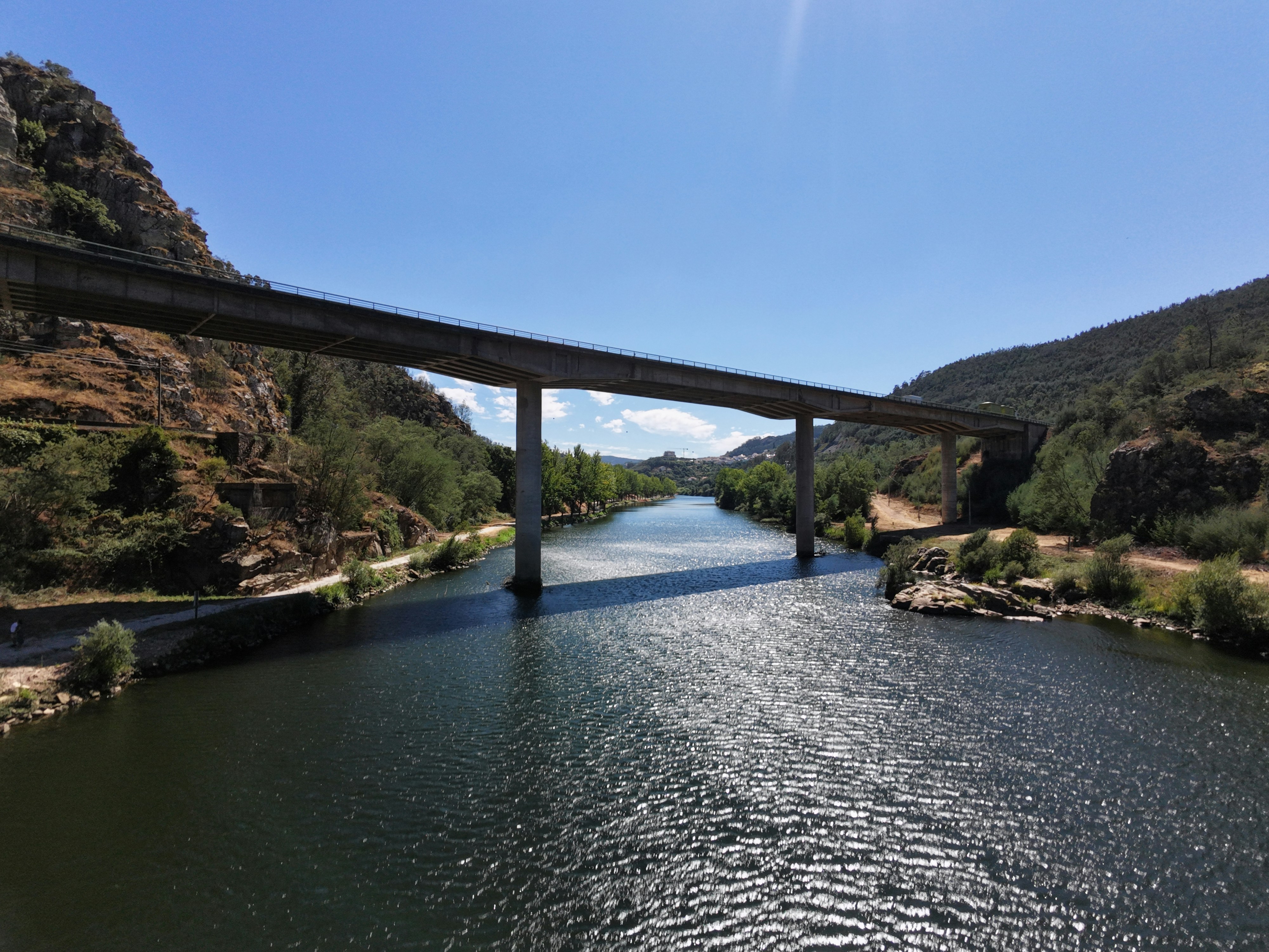 A concrete bridge crosses a river between rocky hills with trees and paths along the banks.