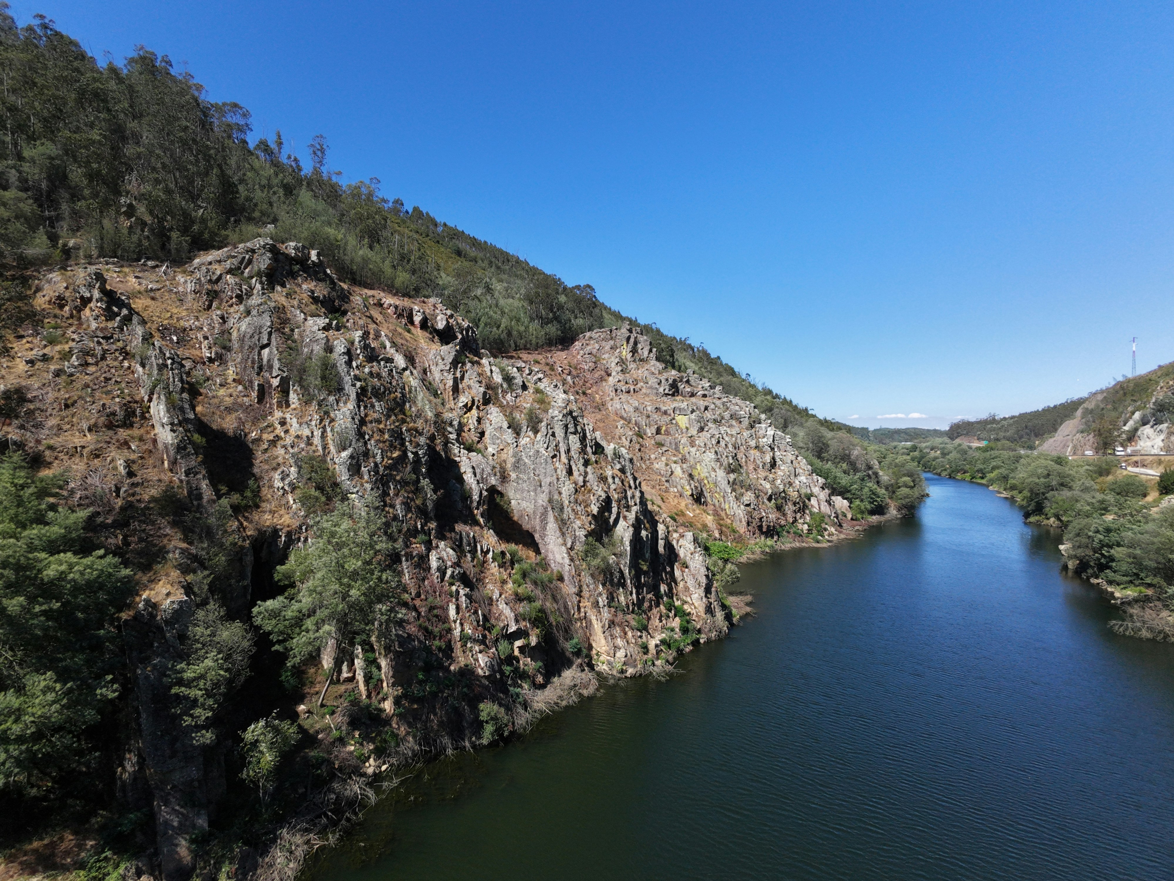 A river runs beside rocky cliffs and wooded hills, with a road along the opposite bank.