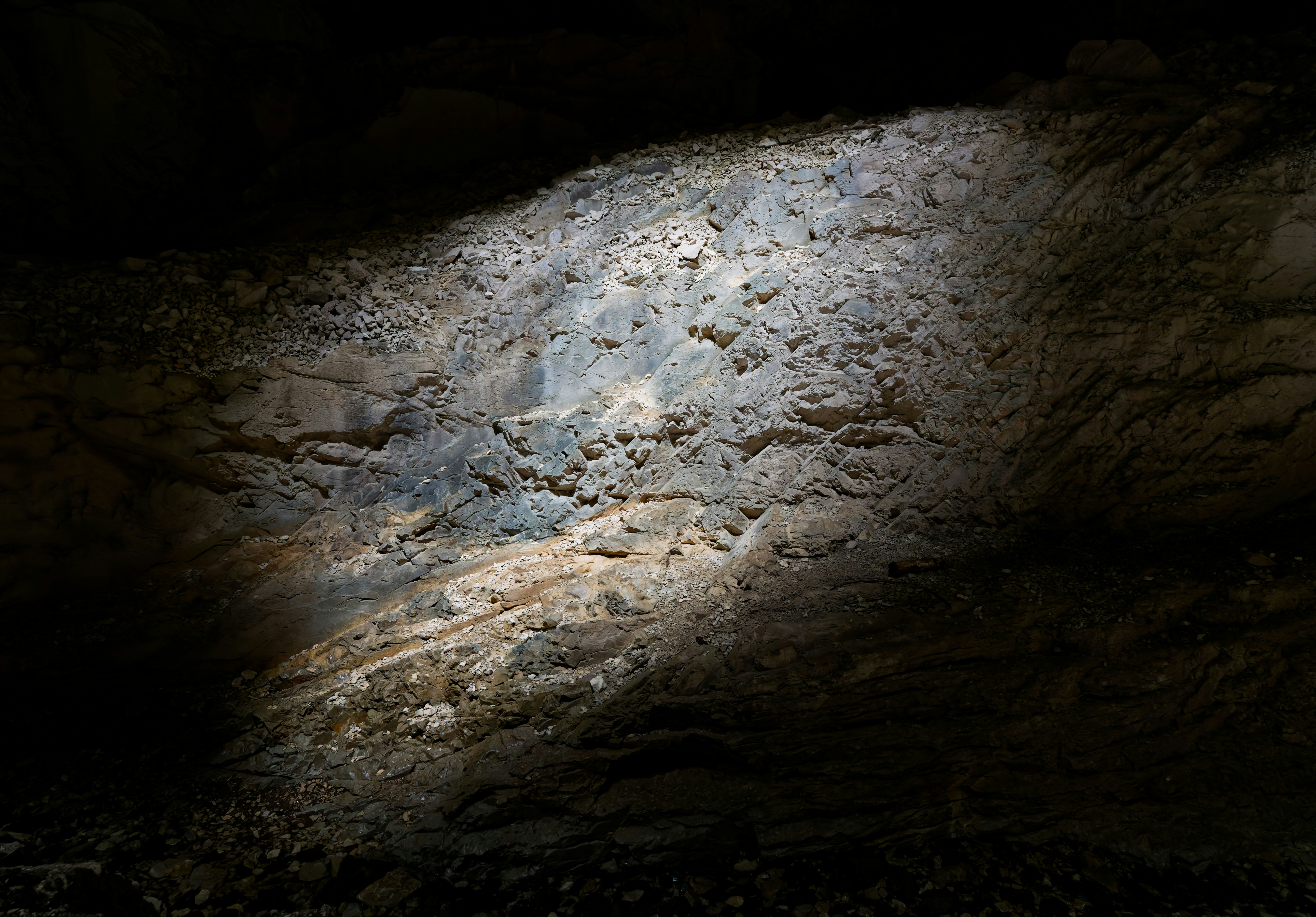 Rock wall illuminated by a narrow beam of light revealing textured mineral strata; the scene emphasizes rugged geology and drama within a dark cave setting.