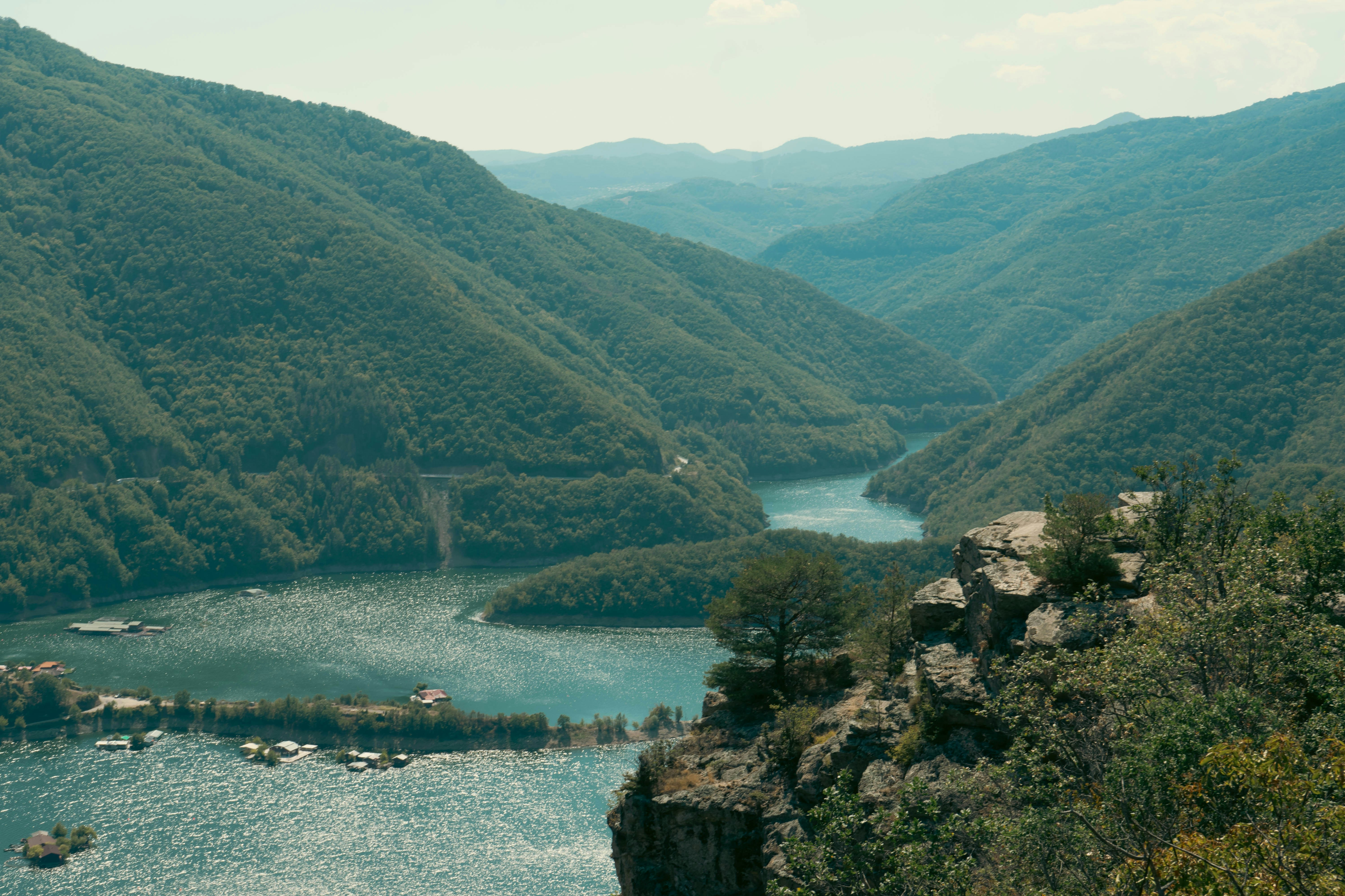 Sunlit river winds through verdant carved valleys with rocky outcrop in the foreground, conveying a sense of depth and serenity. A broad landscape scene suitable for printing as a nature backdrop.
