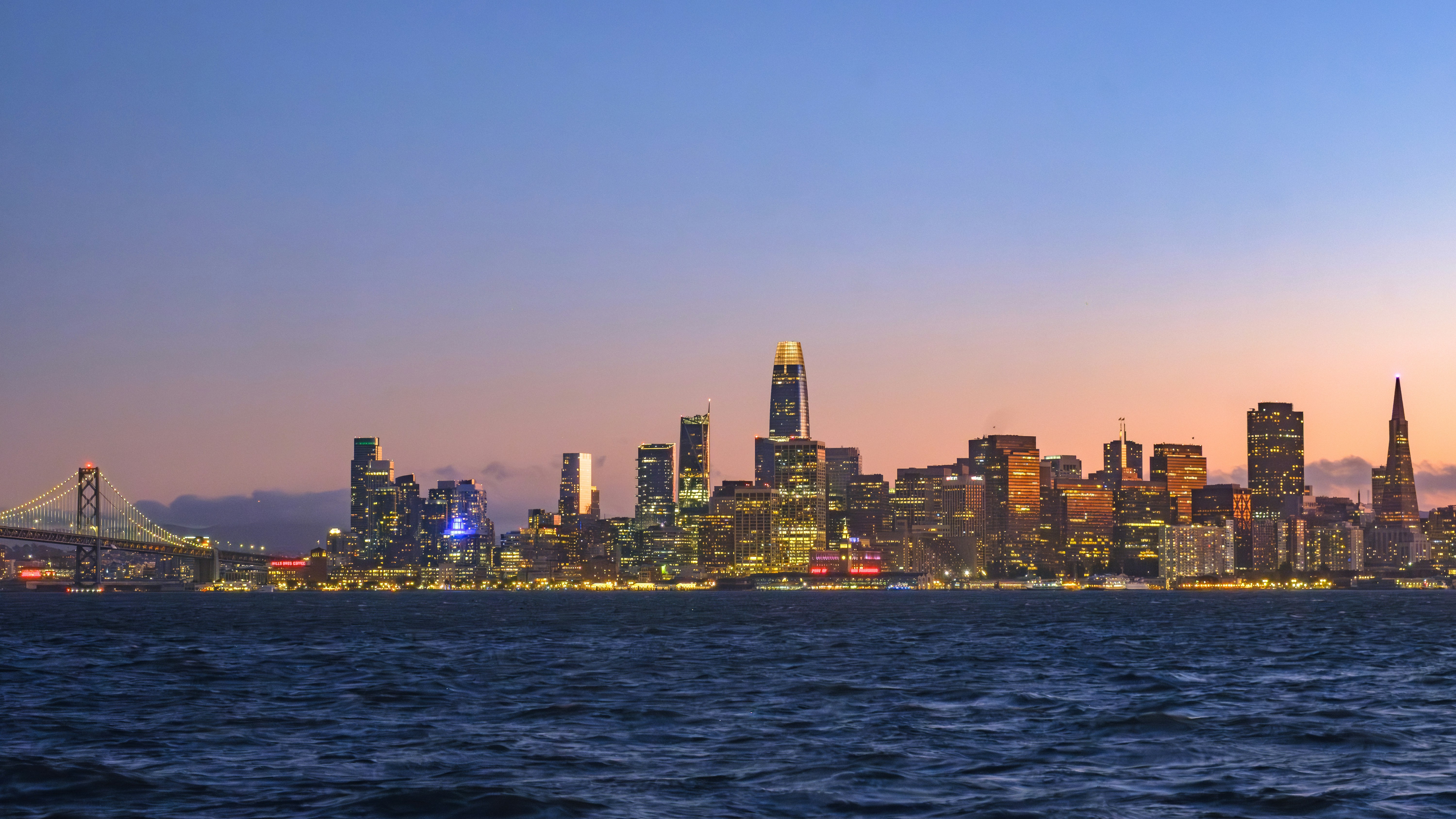 Dusk-lit bay with a vibrant city skyline along the horizon, water in the foreground and silhouettes of skyscrapers shimmering with lights.