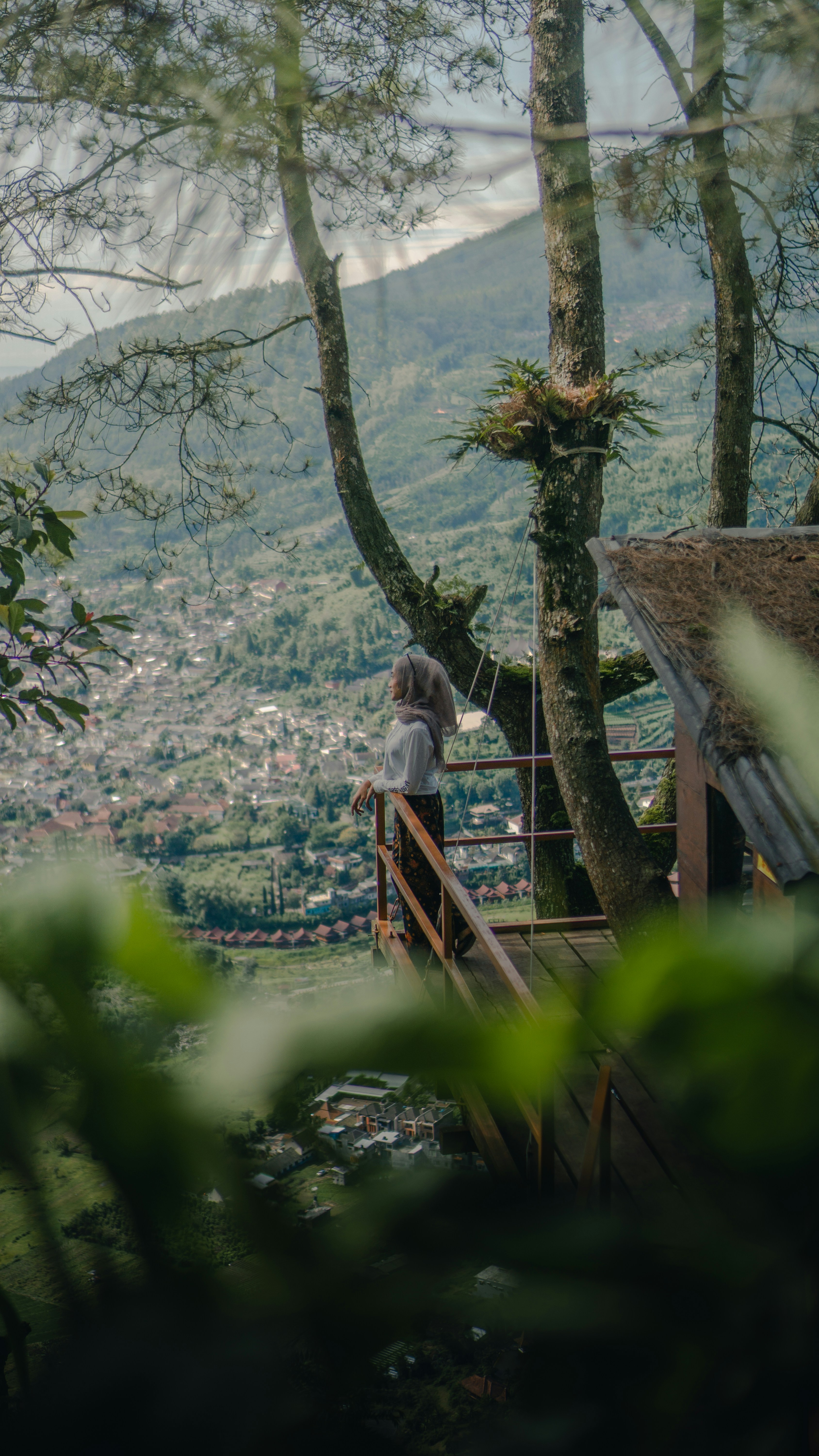 Woman stands on a wooden balcony amid trees, gazing toward a valley town below; the scene is framed by branches and a misty hillside, with a small shelter partially visible. This is a photography of a real scene.