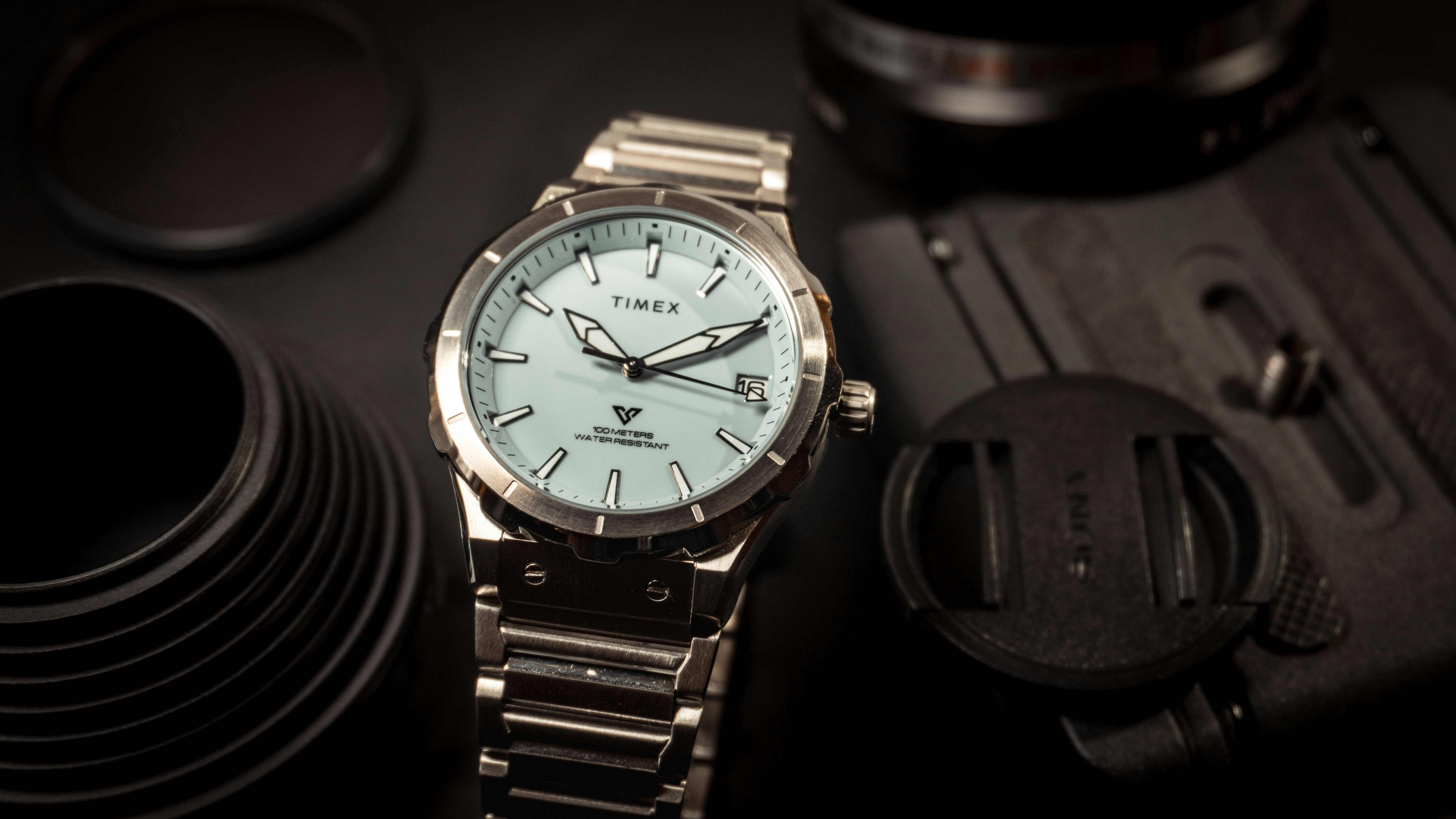 Close-up of a silver Timex wristwatch resting among camera accessories and lenses, highlighting craftsmanship and metallic details.