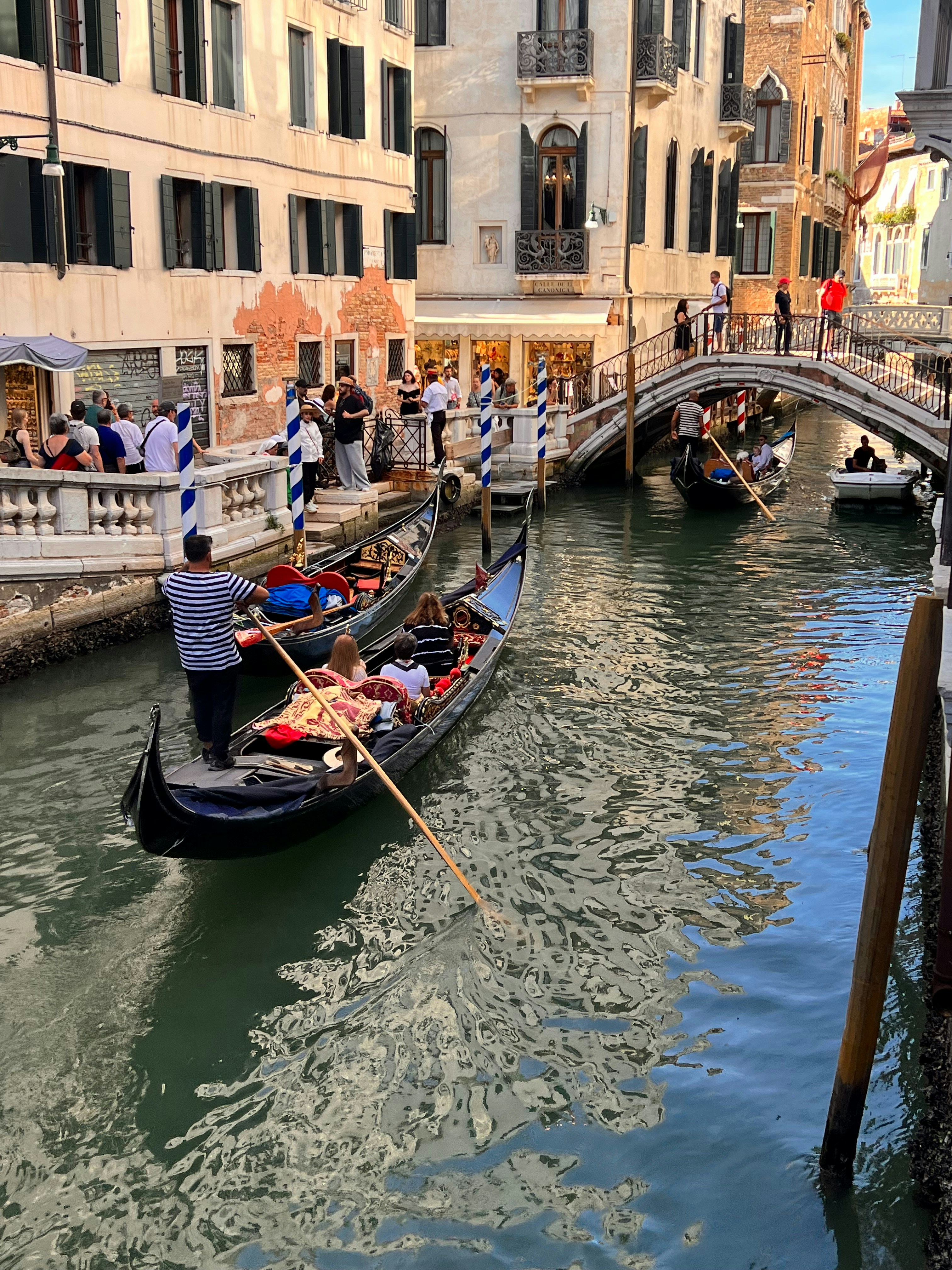 Vibrant canal scene with gondolas and people along a stone bridge, classic Venetian architecture lining the water's edge, and calm reflections on the canal surface. A lively urban moment captured along a narrow waterway.