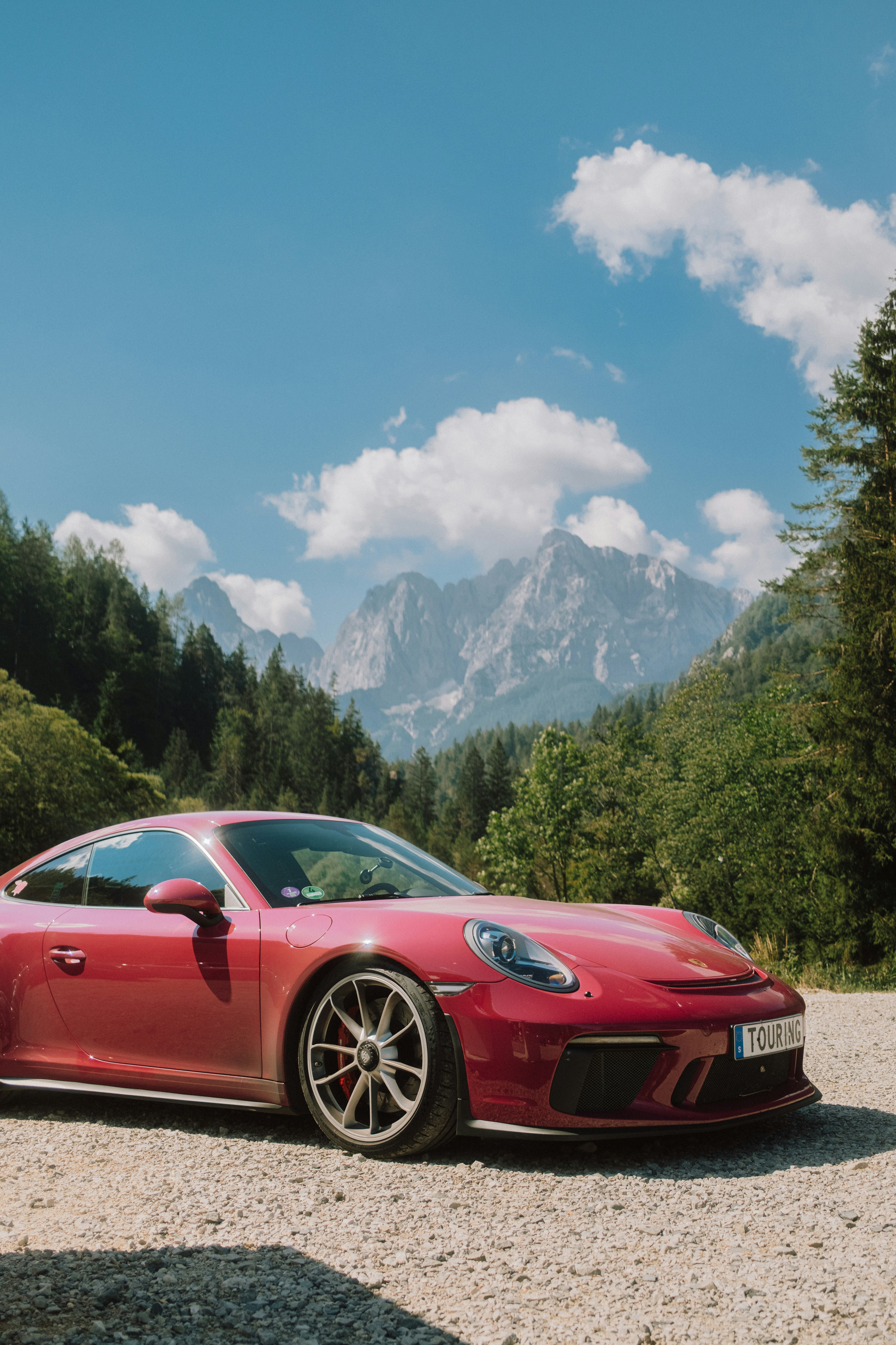 Crimson Porsche parked on a gravel lot with a backdrop of evergreen trees and snow-capped mountains under a bright blue sky.
