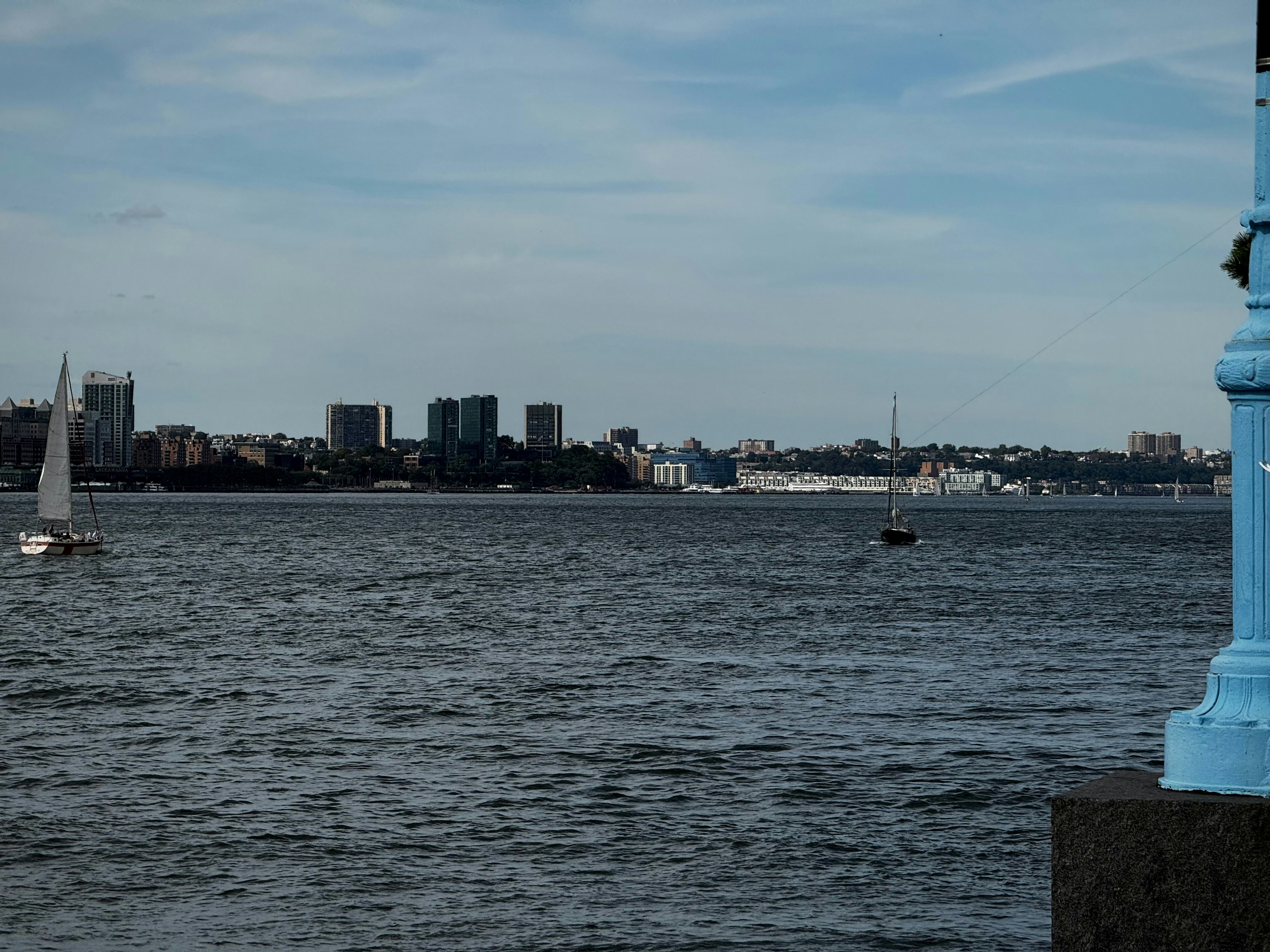 Calm water with a distant city skyline across the bay and a blue lamppost on the right; a sailboat and a small craft drift under a clear sky.