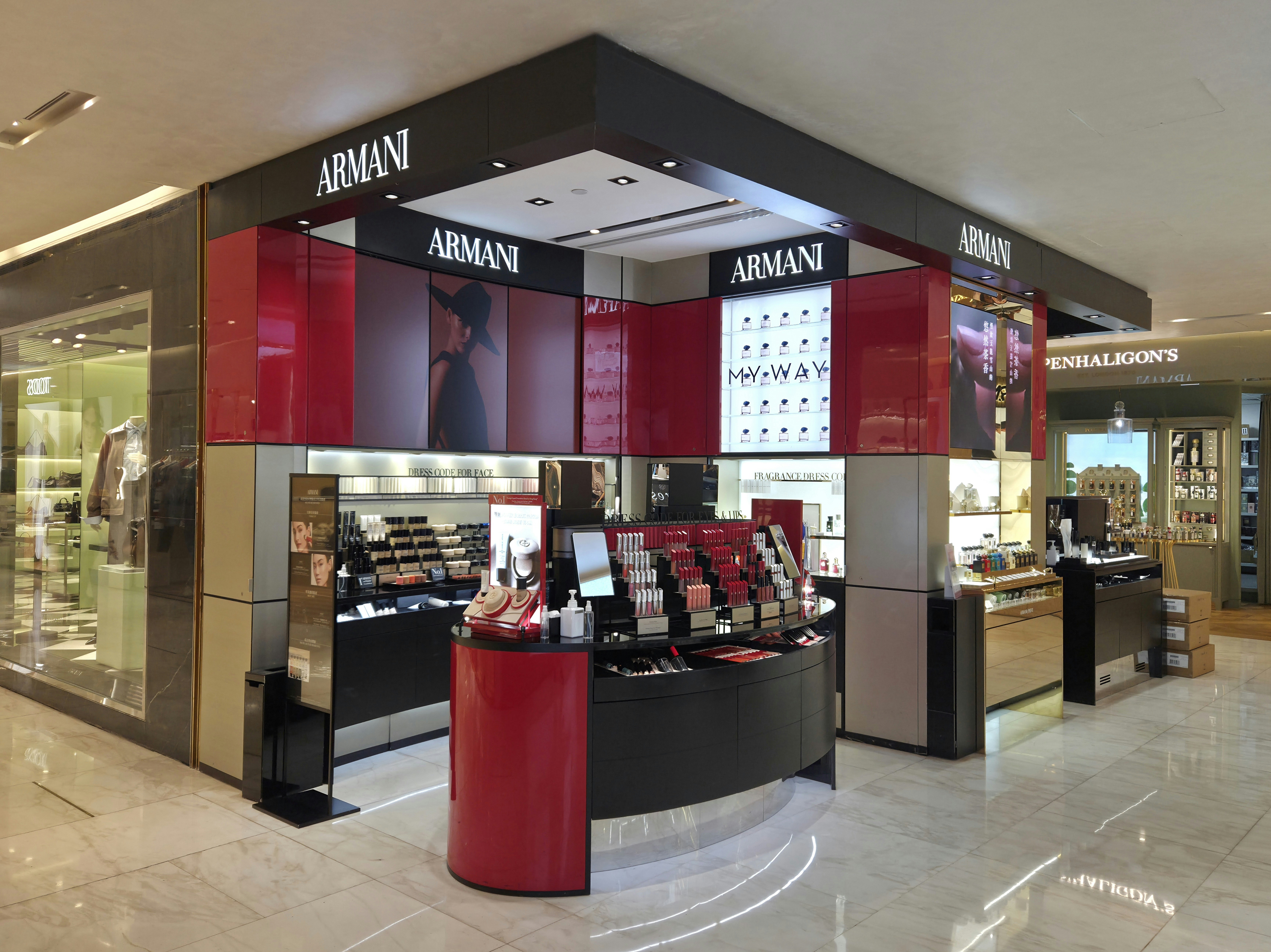 Armani storefront inside a mall, featuring a rounded red display table and illuminated product shelves. The scene highlights premium accessories and a modern retail environment.