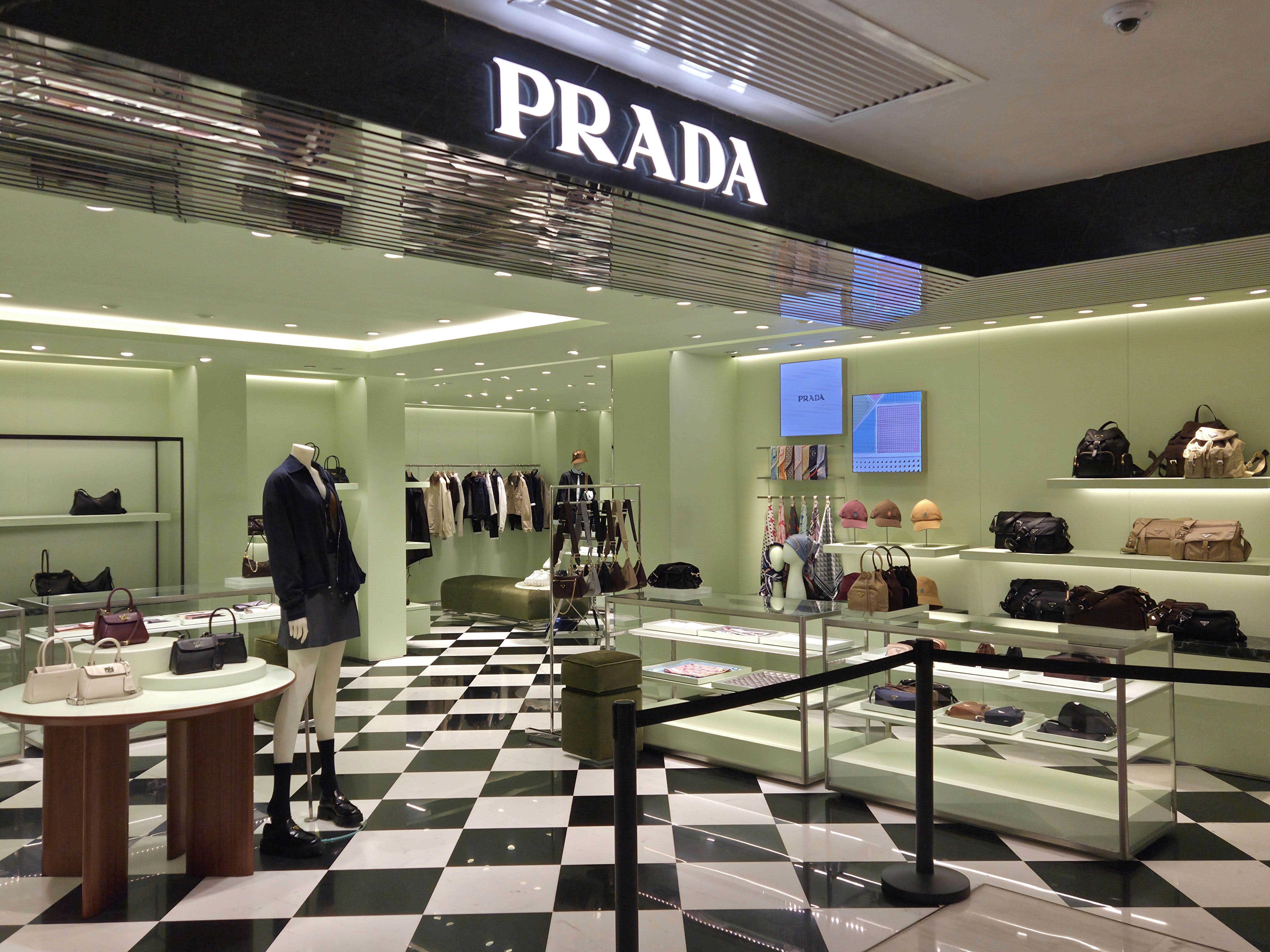 End-to-end view of a Prada storefront interior with mint-green walls, a black-and-white checkered floor, and glass display cases filled with bags and accessories.