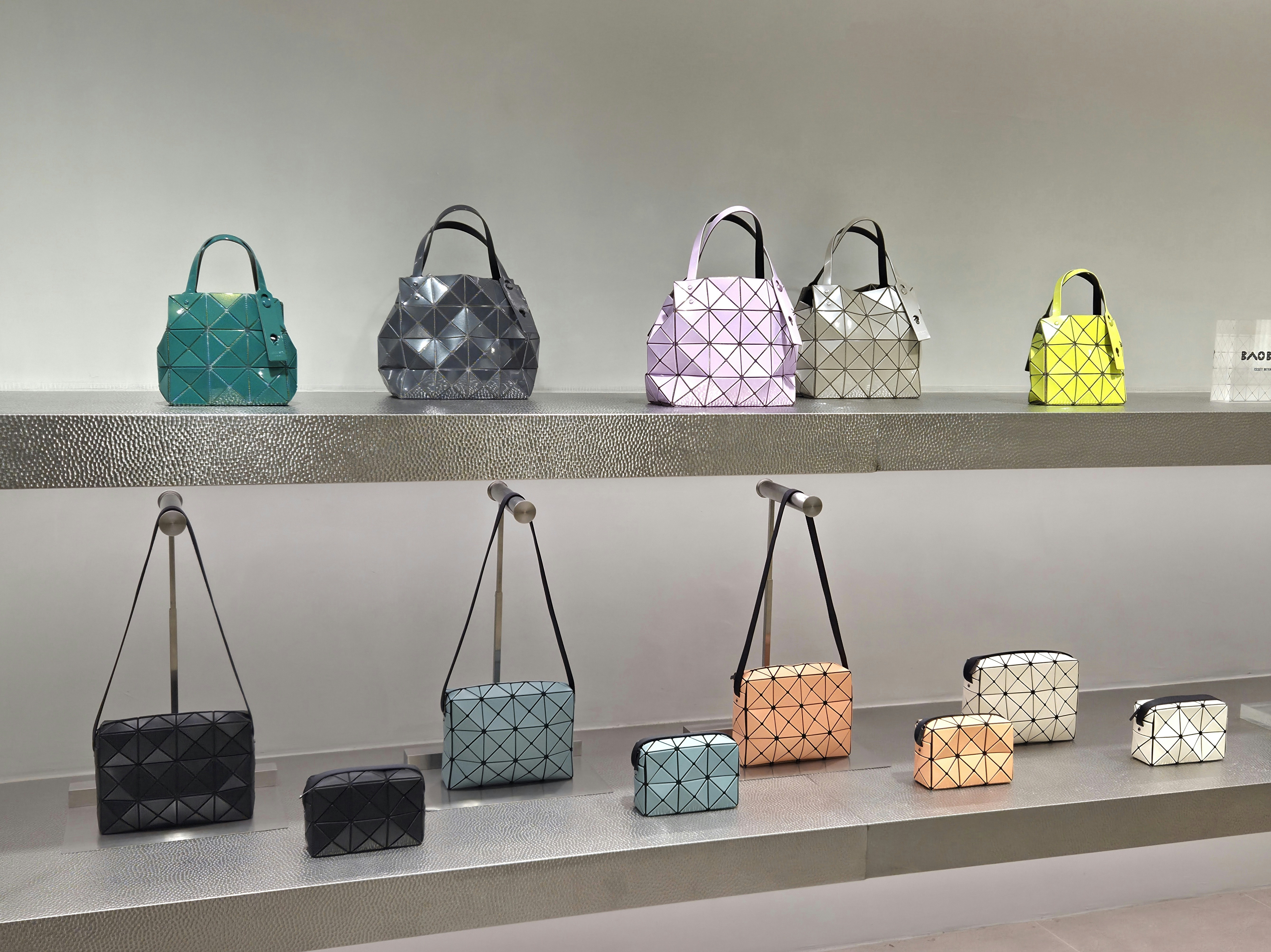 A display of geometric, faceted handbags in various colors arranged on two illuminated shelves in a storefront. The collection features small and medium purses with triangle-panel surfaces, suggesting a designer line and a glossy, modern aesthetic.