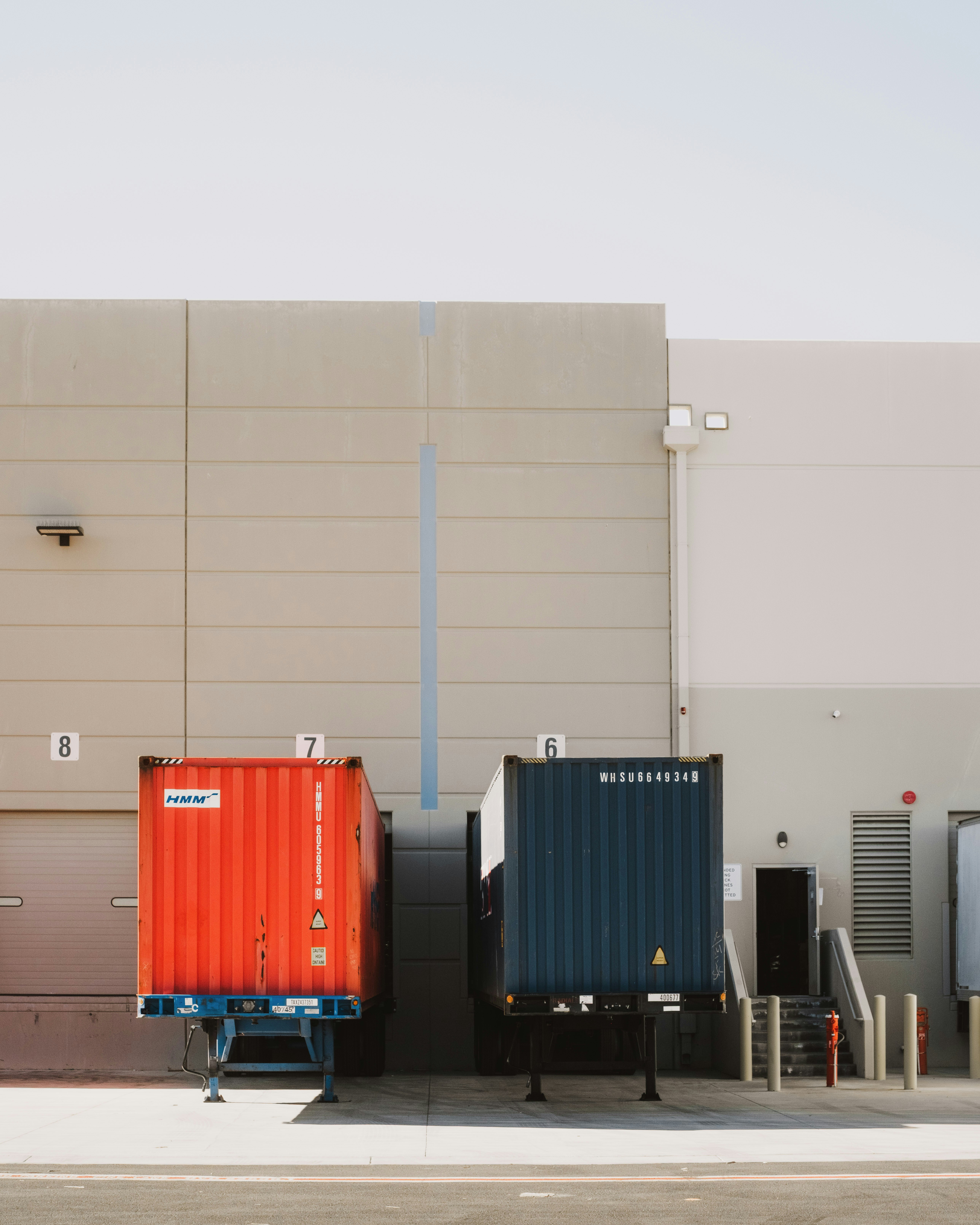 Two cargo containers sit side by side in front of a warehouse, with a pale, linear backdrop and a clear sky above.