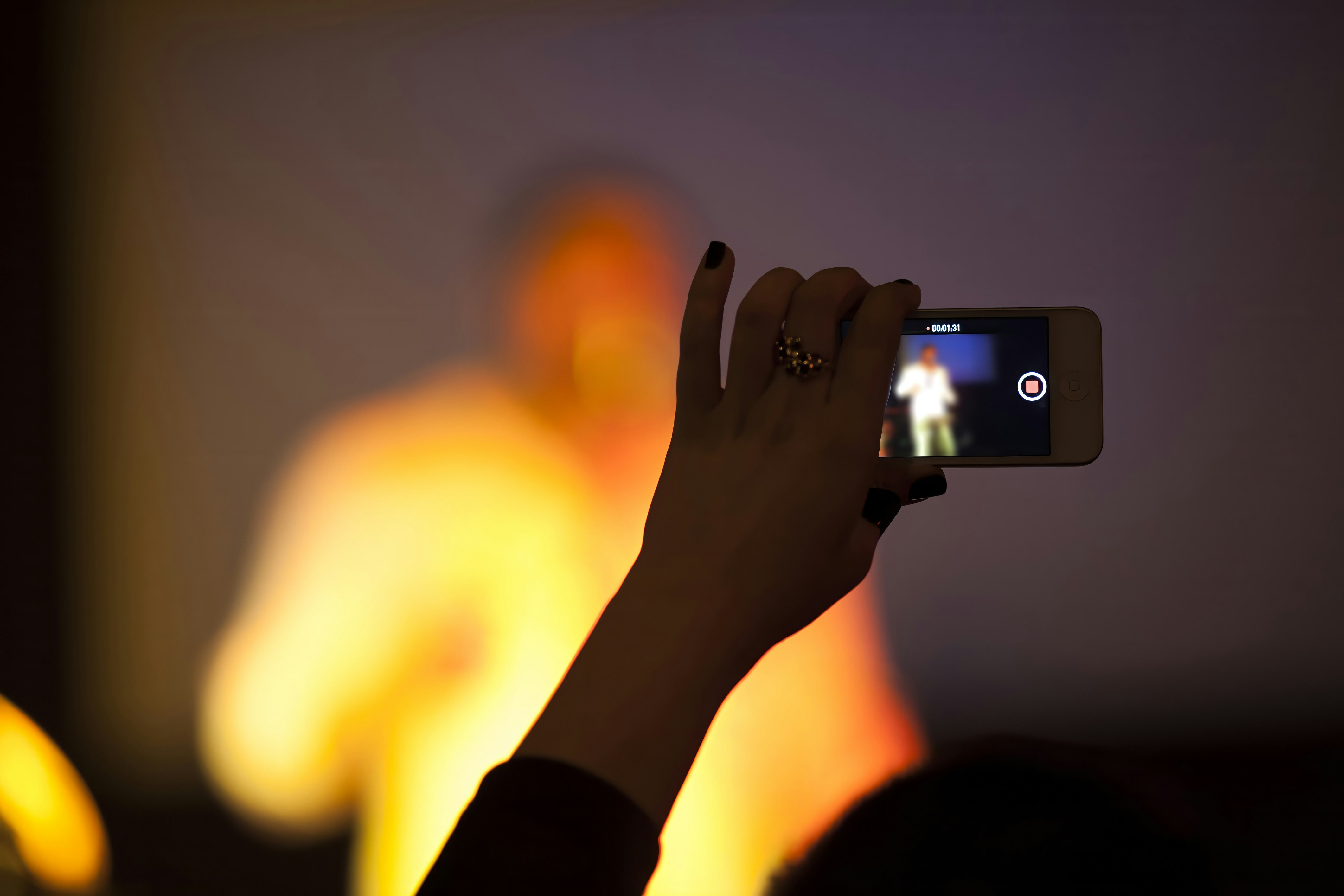 A blurred stage scene is observed through a raised smartphone aimed at the performance, with the phone screen showing a clearer subject. A logo or device screen hints at a live event; the image emphasizes user-held capture amid a warm, amber-lit background.