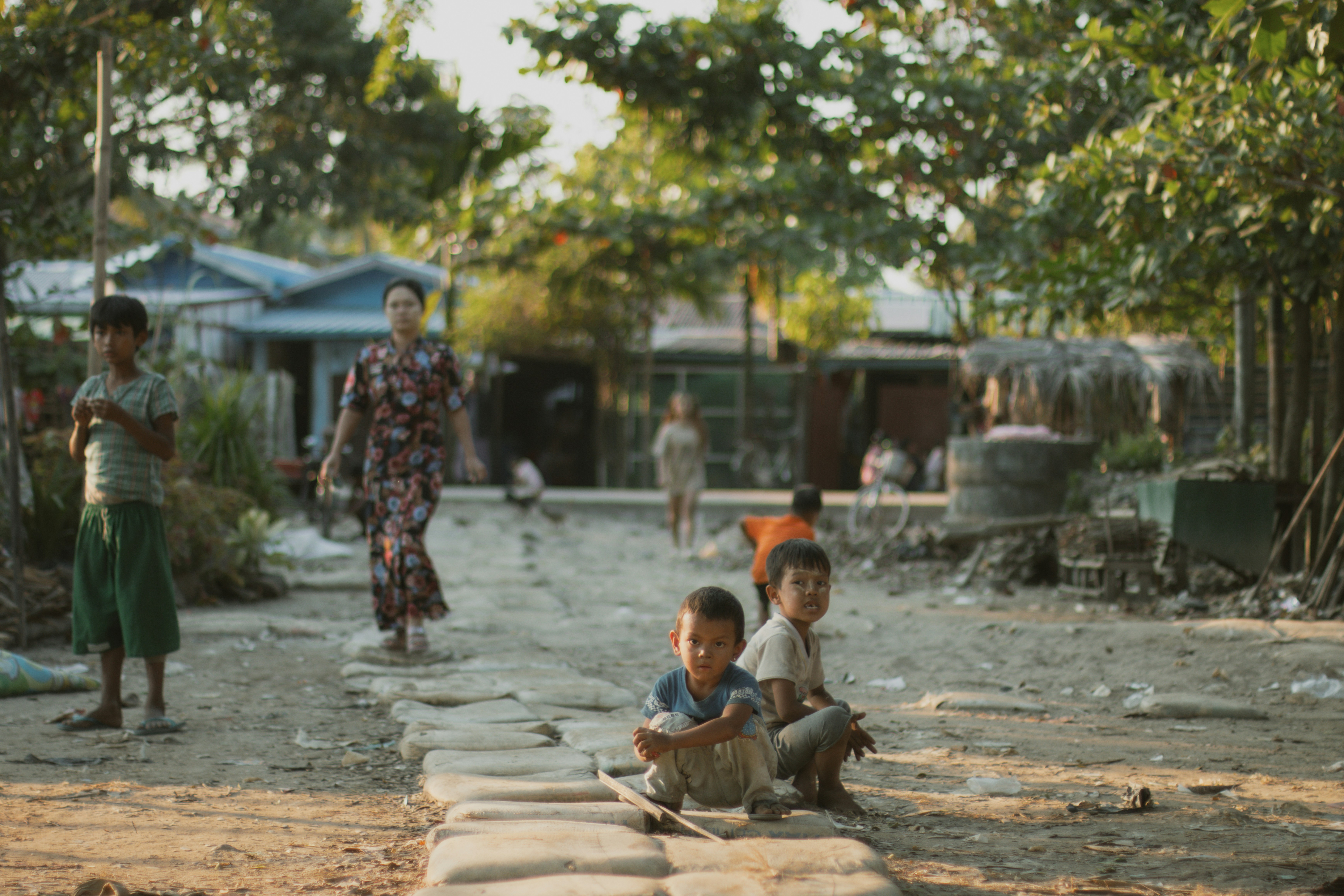 Two children pause on a sunlit dirt path as a village settles into the afternoon