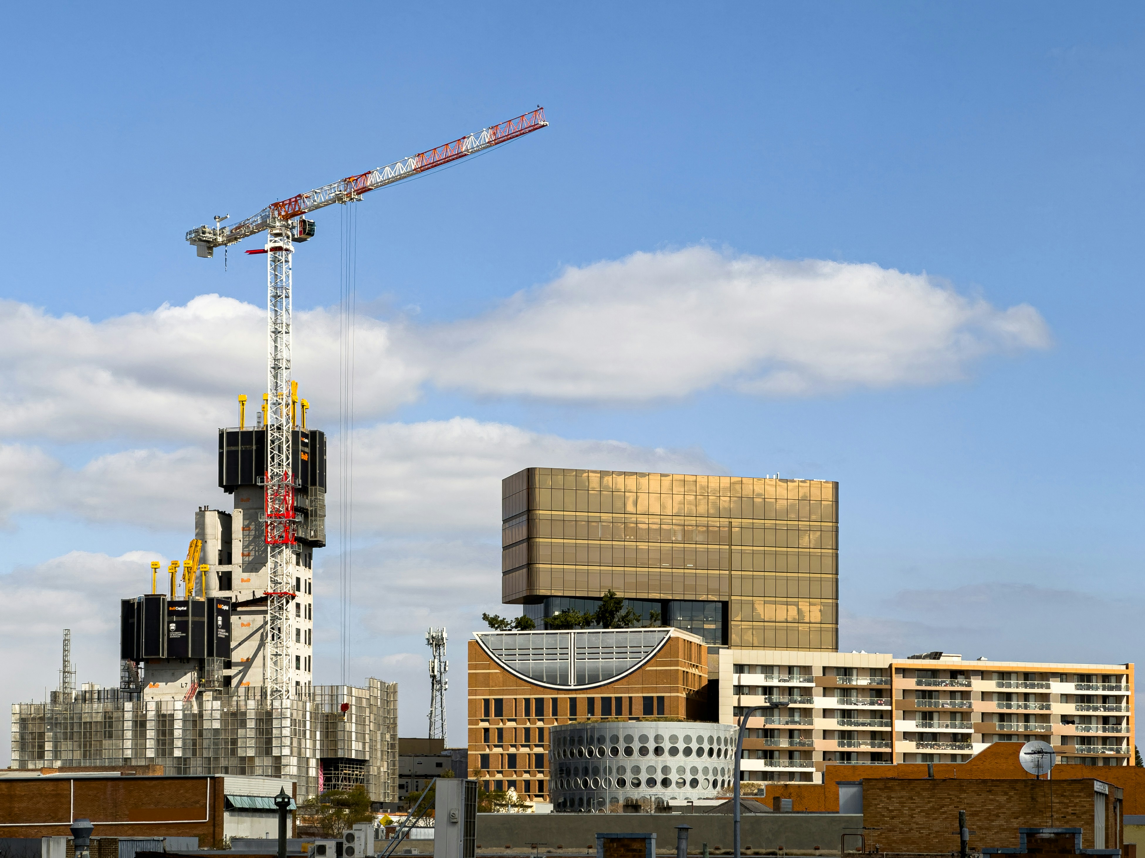 Construction cranes loom beside a modern, gold-toned building cluster, capturing a daytime urban scene with blue sky and white clouds.