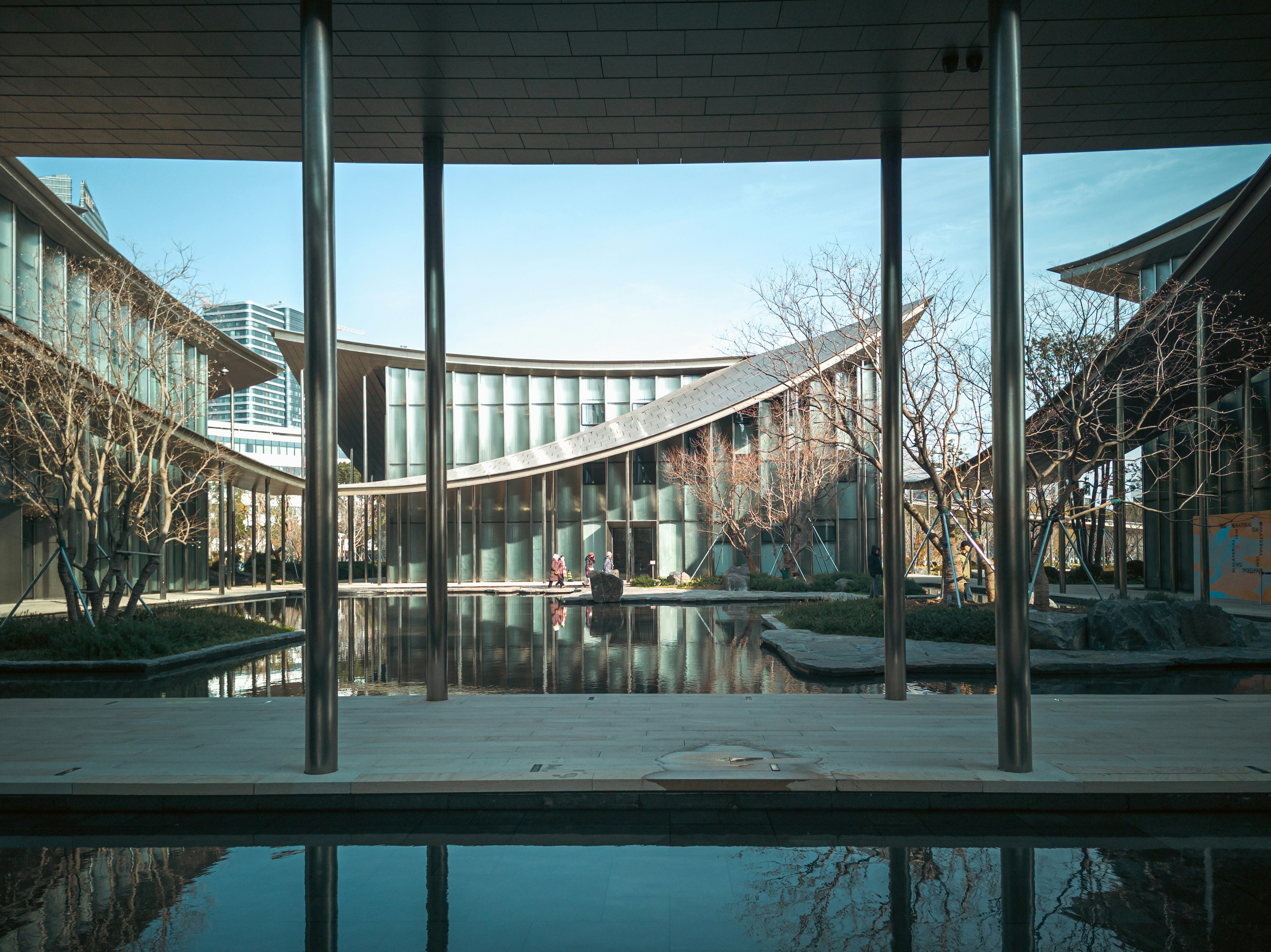 Modern glass buildings with curved roofs surrounding a reflective pool in a courtyard
