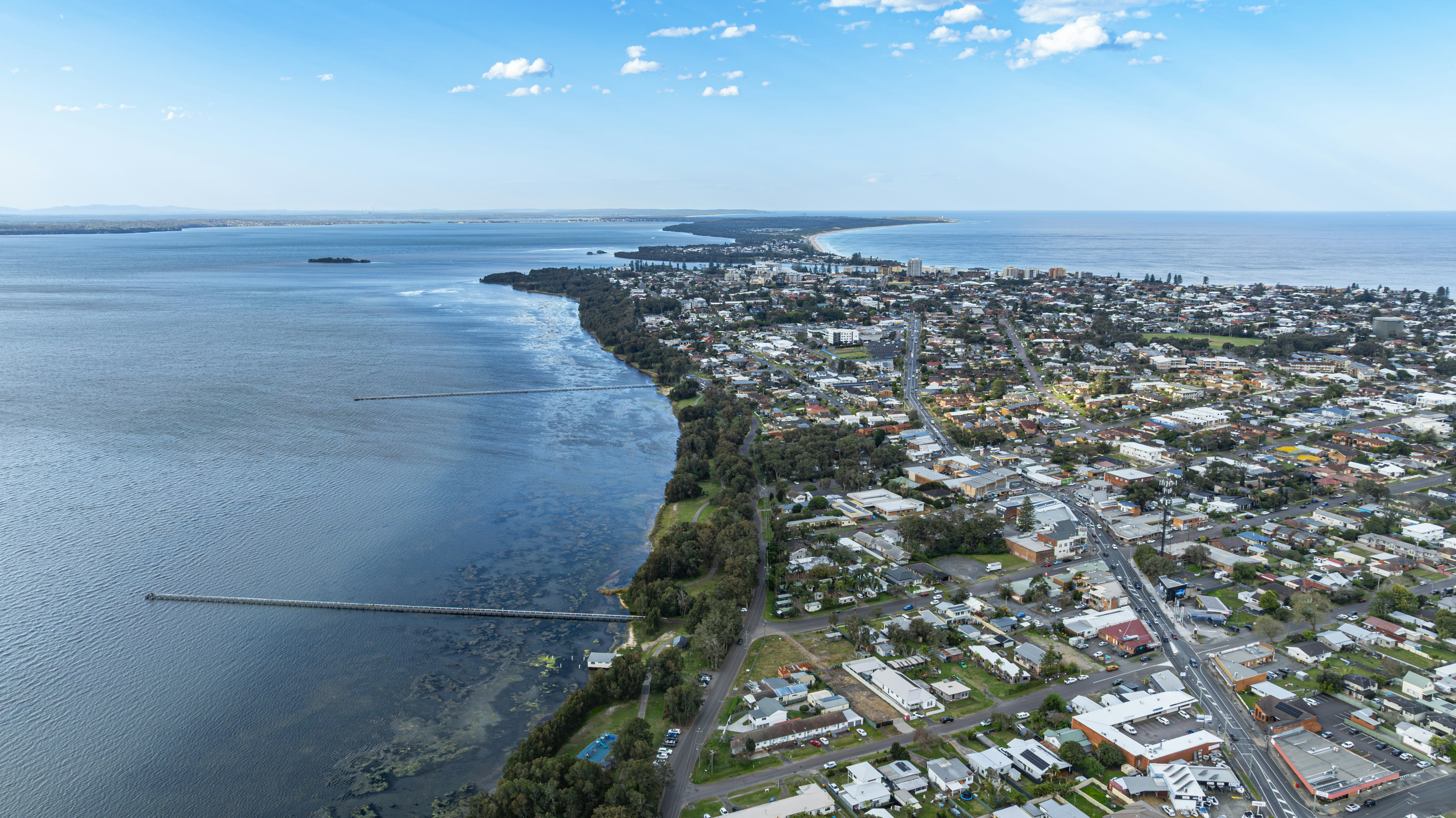 Aerial view of a coastal suburb with houses and streets lining the shoreline; the water meets land along a curved coast, with a breakwater extending into the sea. A logo or branding element is not visible; overall scene is a real-world geographic capture.