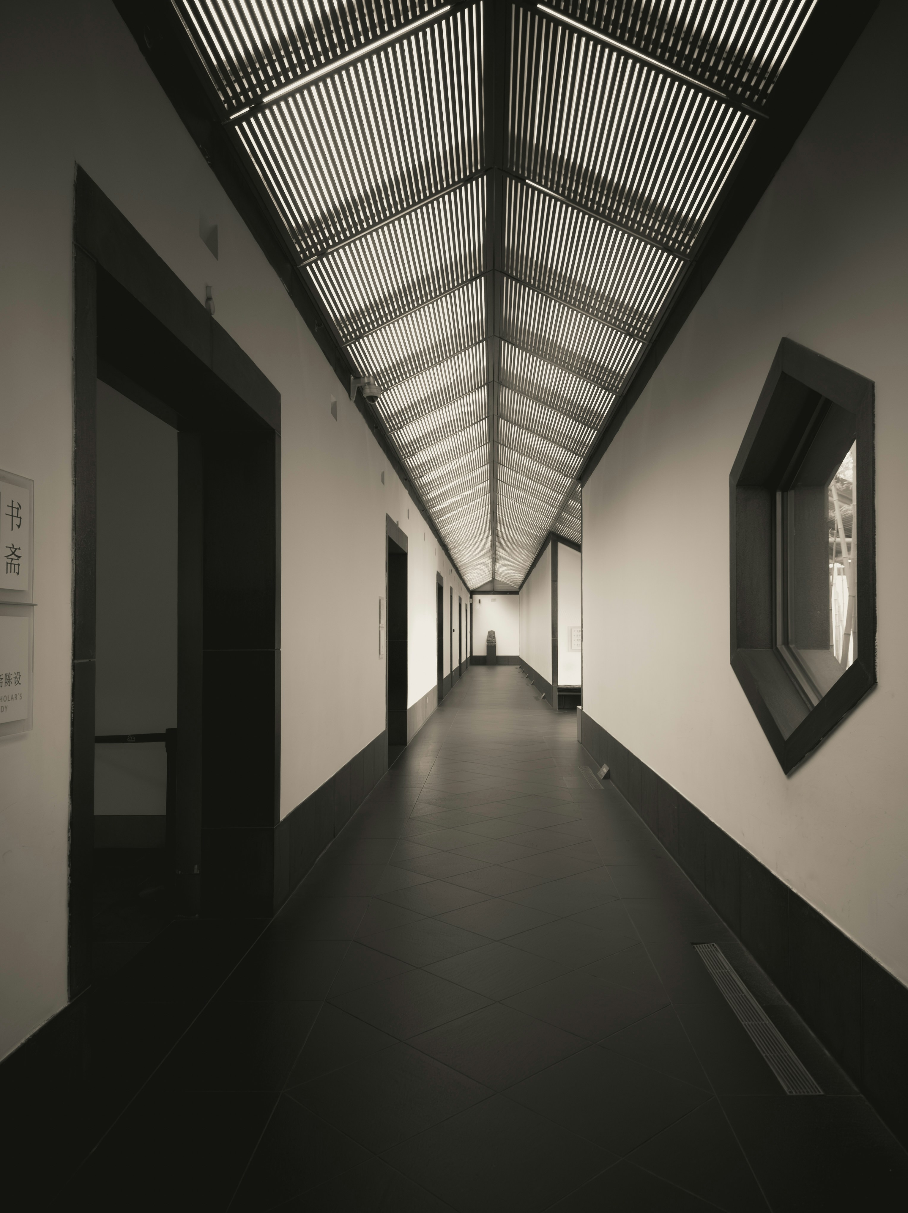 A long corridor with white walls, dark floors, and a slatted glass ceiling
