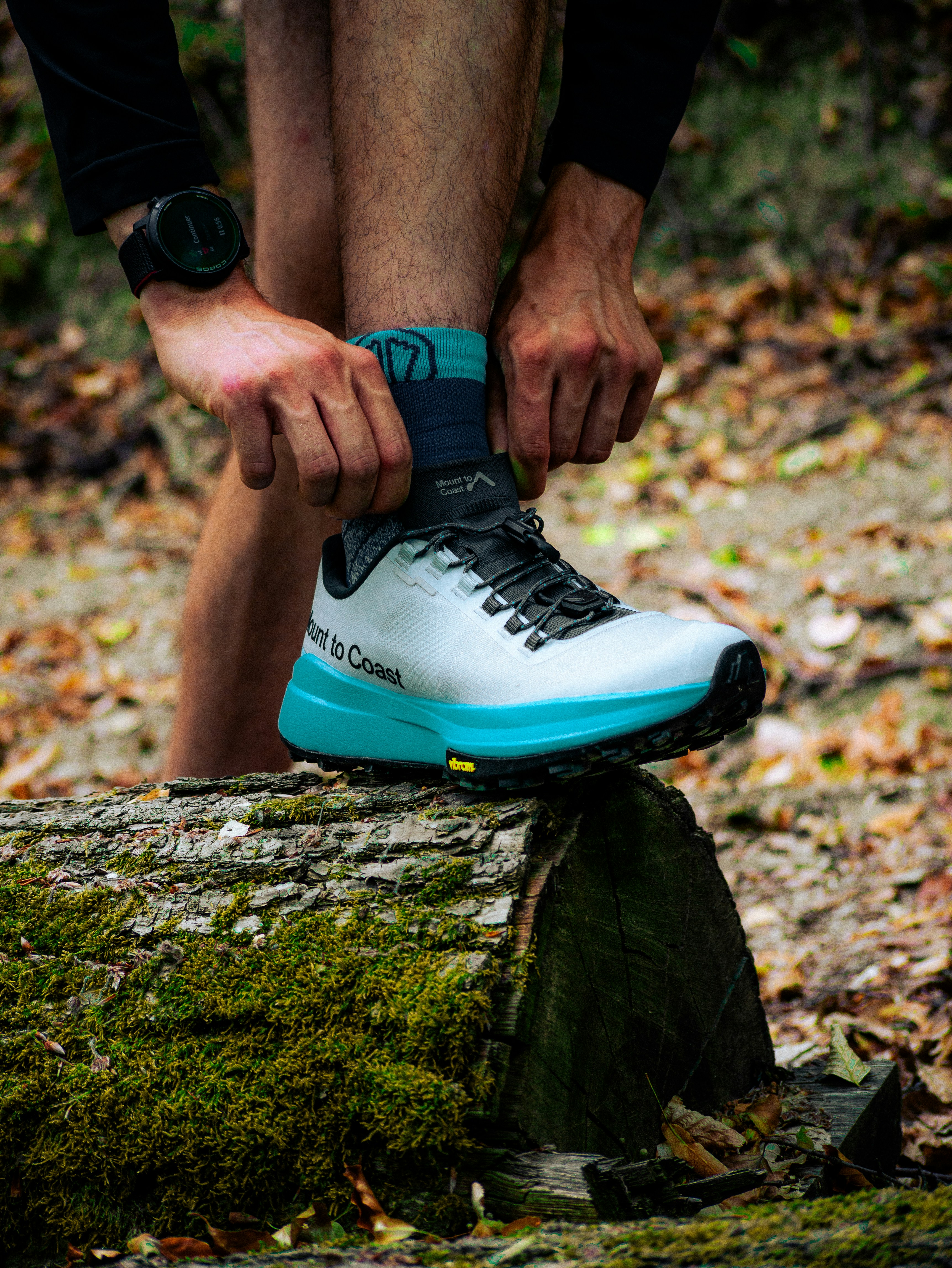 Runner tightens the laces of a white-and-teal trail shoe as they stand on a mossy log in a forest floor covered with autumn leaves. A close-up emphasizes the footwear and ankle strap details.