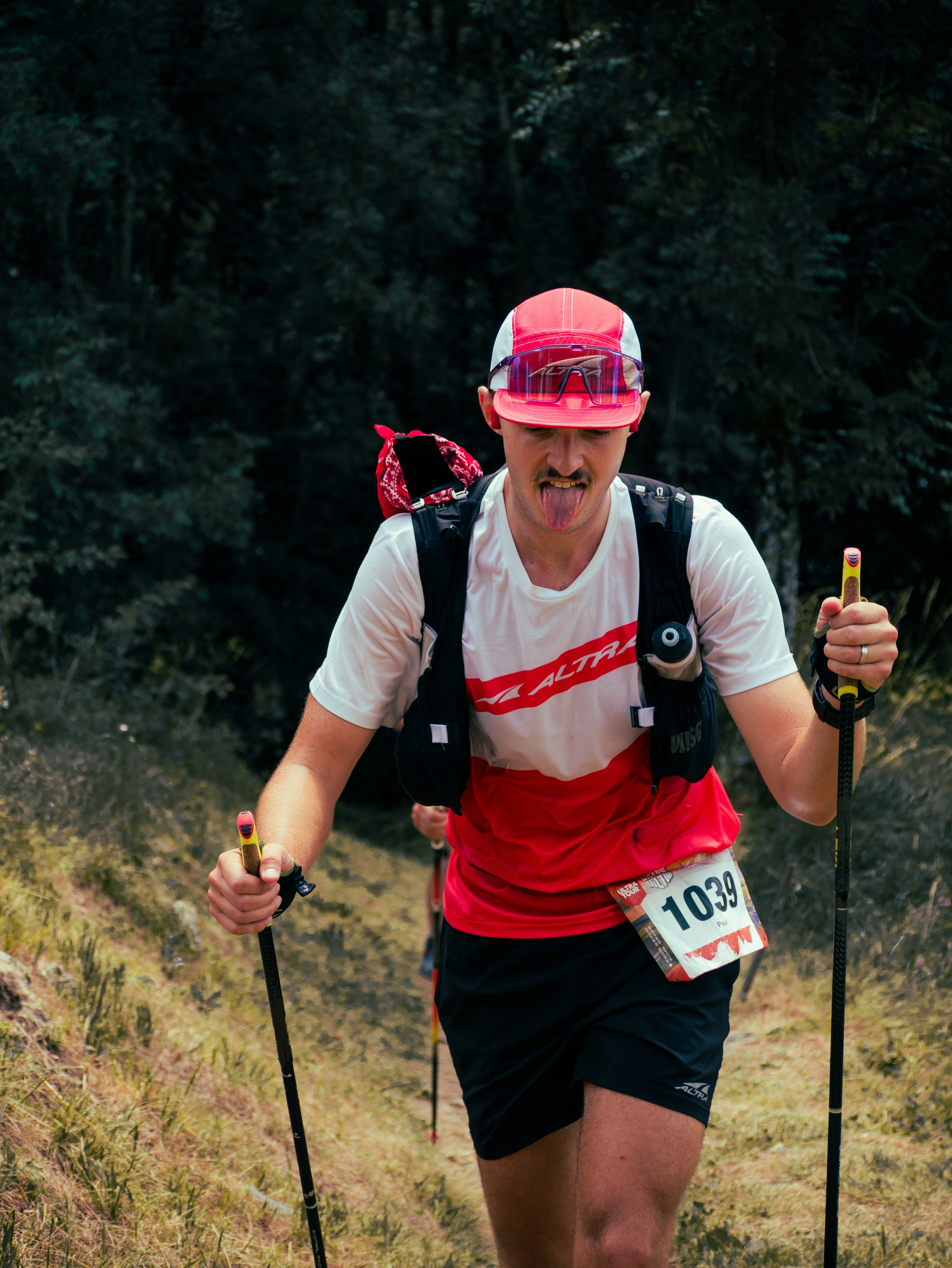 Bold trail runner surges forward with trekking poles in a sunlit dirt path, wearing a red and white kit and a race bib; a backpack and helmet-style cap complete the gear.