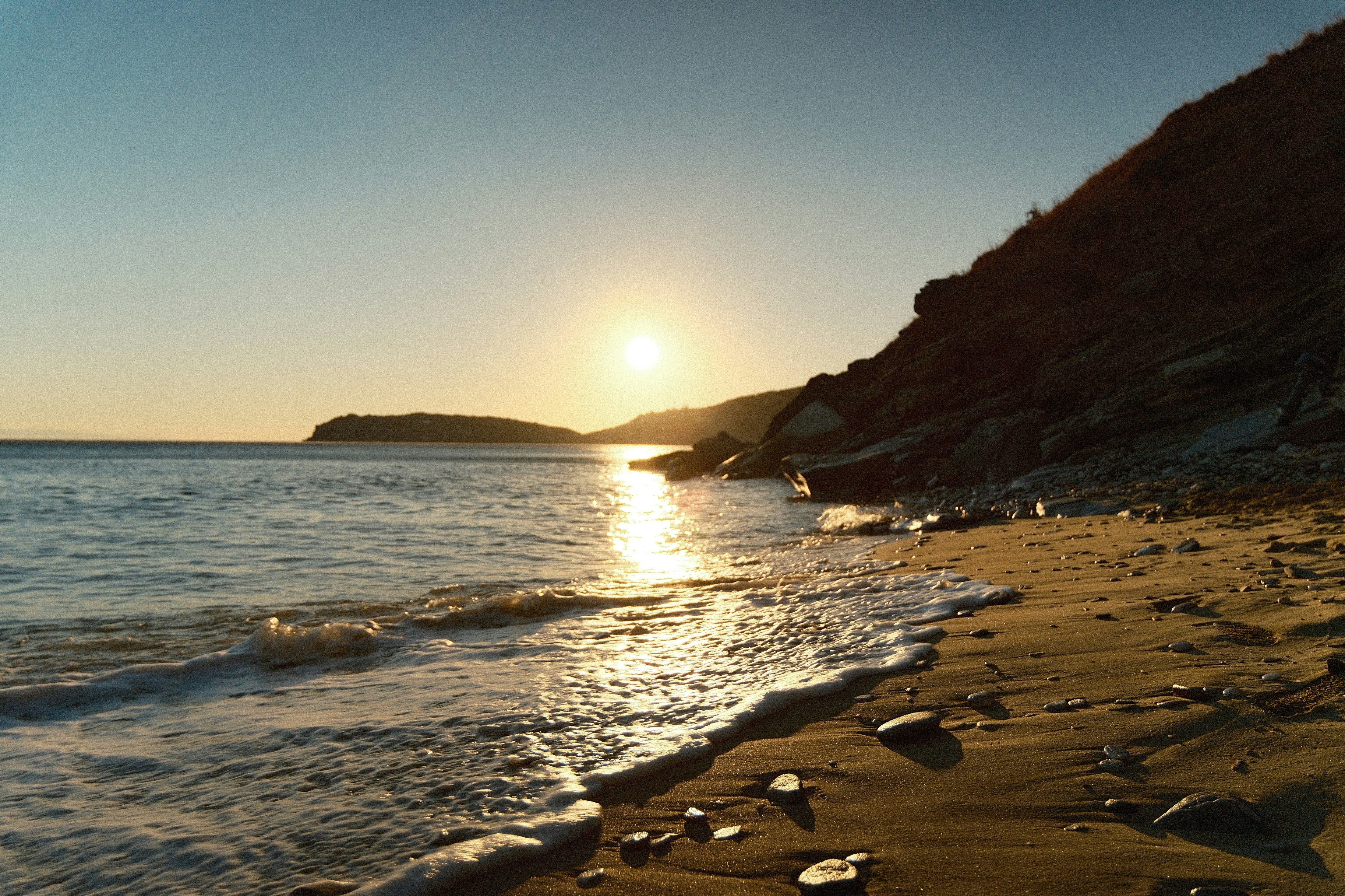 Sunlit shoreline during golden hour with gentle waves washing onto a sandy beach and a sloping hillside on the right; a calm horizon and reflection of the sun on the water. Logo/branding not present; natural landscape.